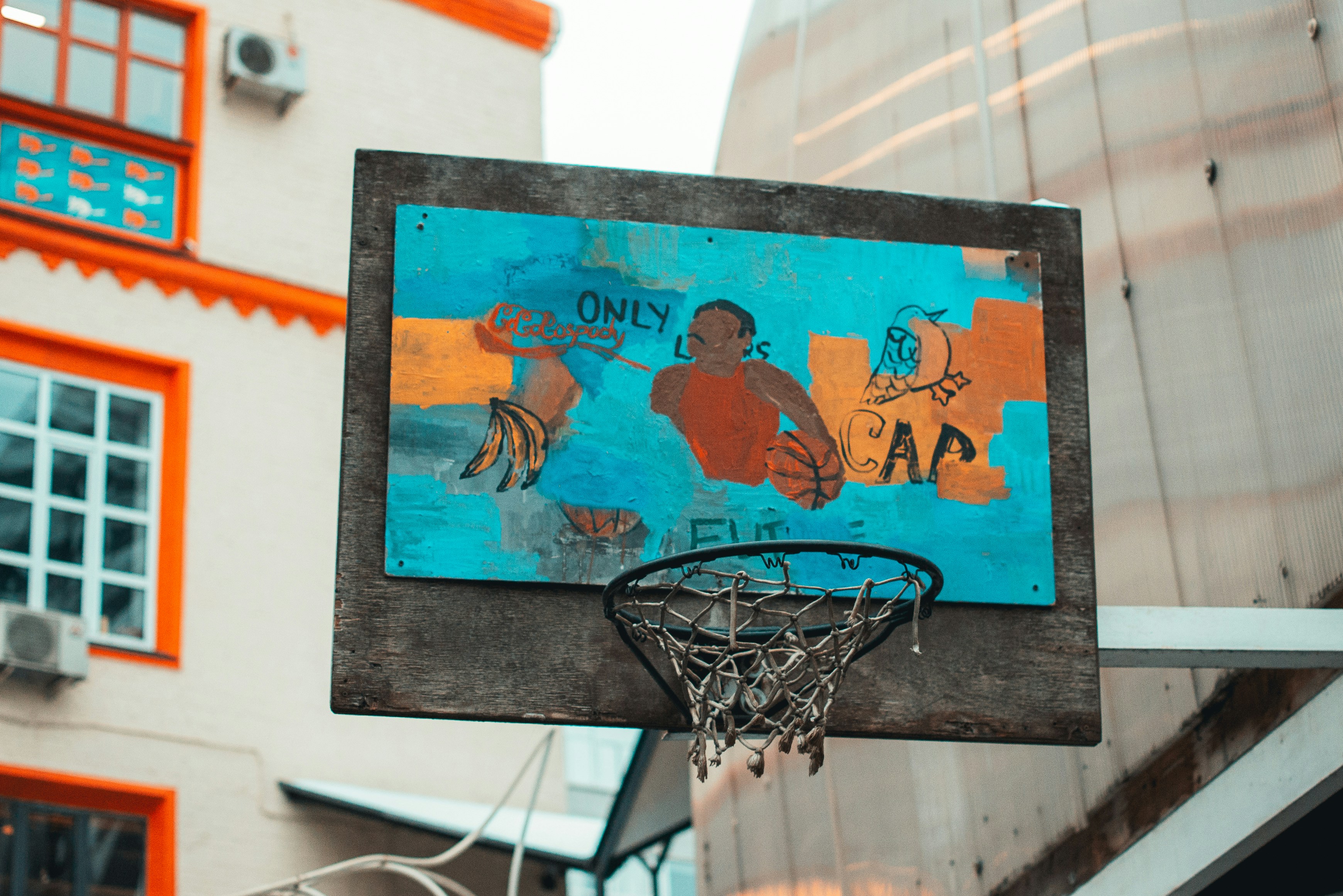 Colorful mural on a basketball backboard featuring playful illustrations and text, set against a backdrop of urban architecture.