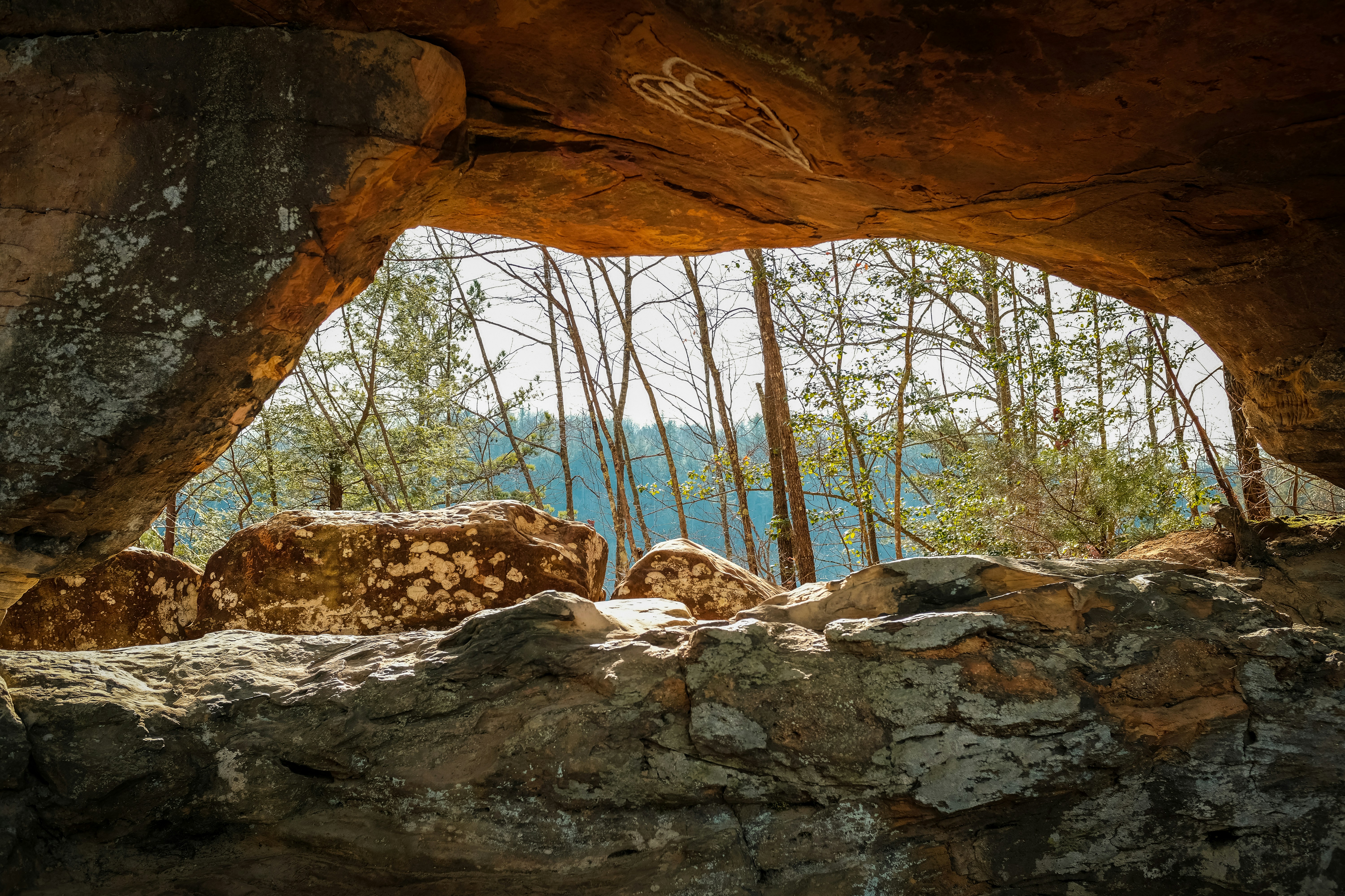 Natural rock formation framing a serene view of trees and distant hills. The sunlight filters through, highlighting the textures of the stone.