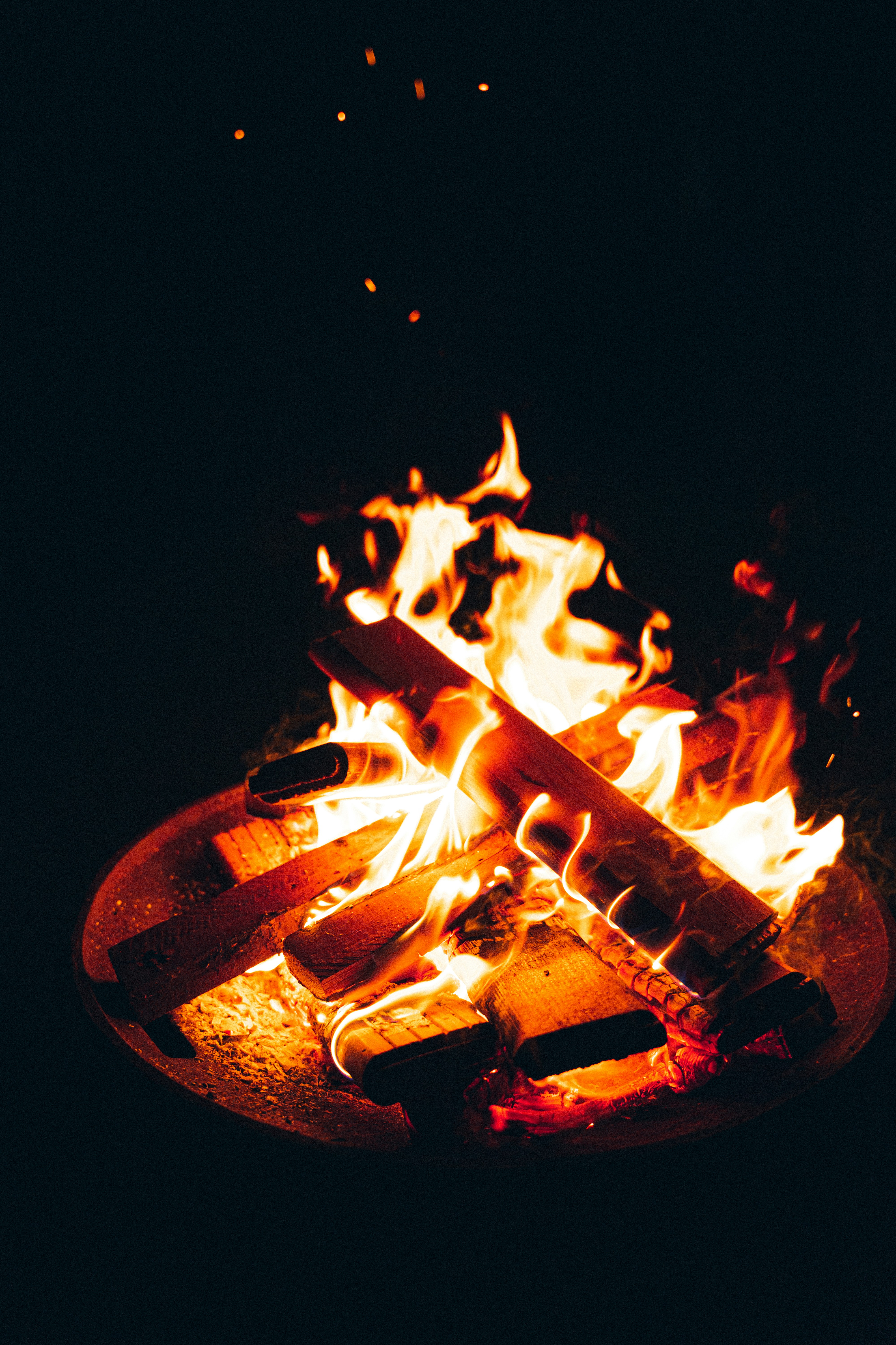 Bright flames dance around logs in a fire pit, illuminating the dark surroundings with a warm glow.