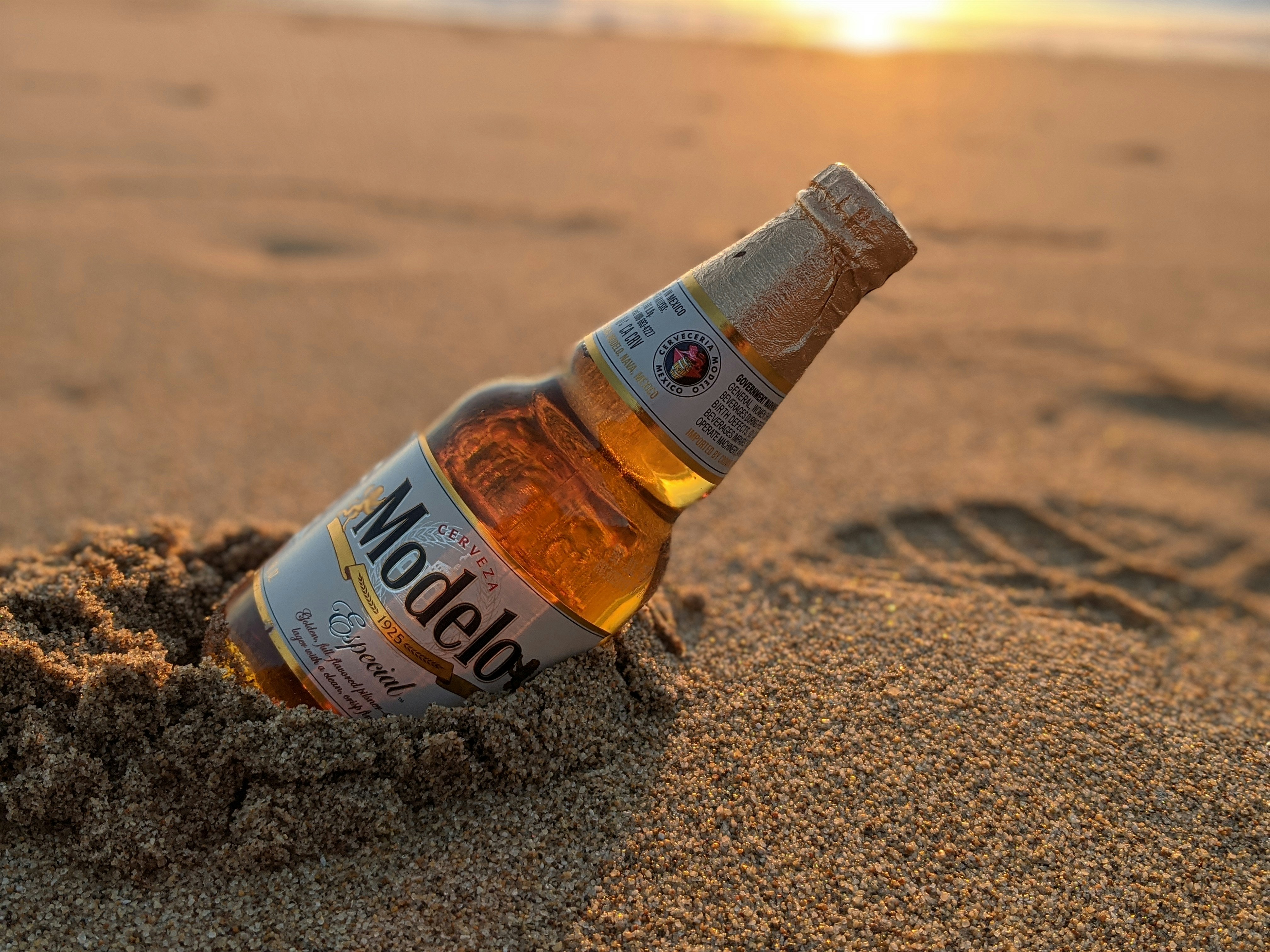 A bottle of Modelo resting in warm sand, illuminated by the soft glow of a setting sun in the background.