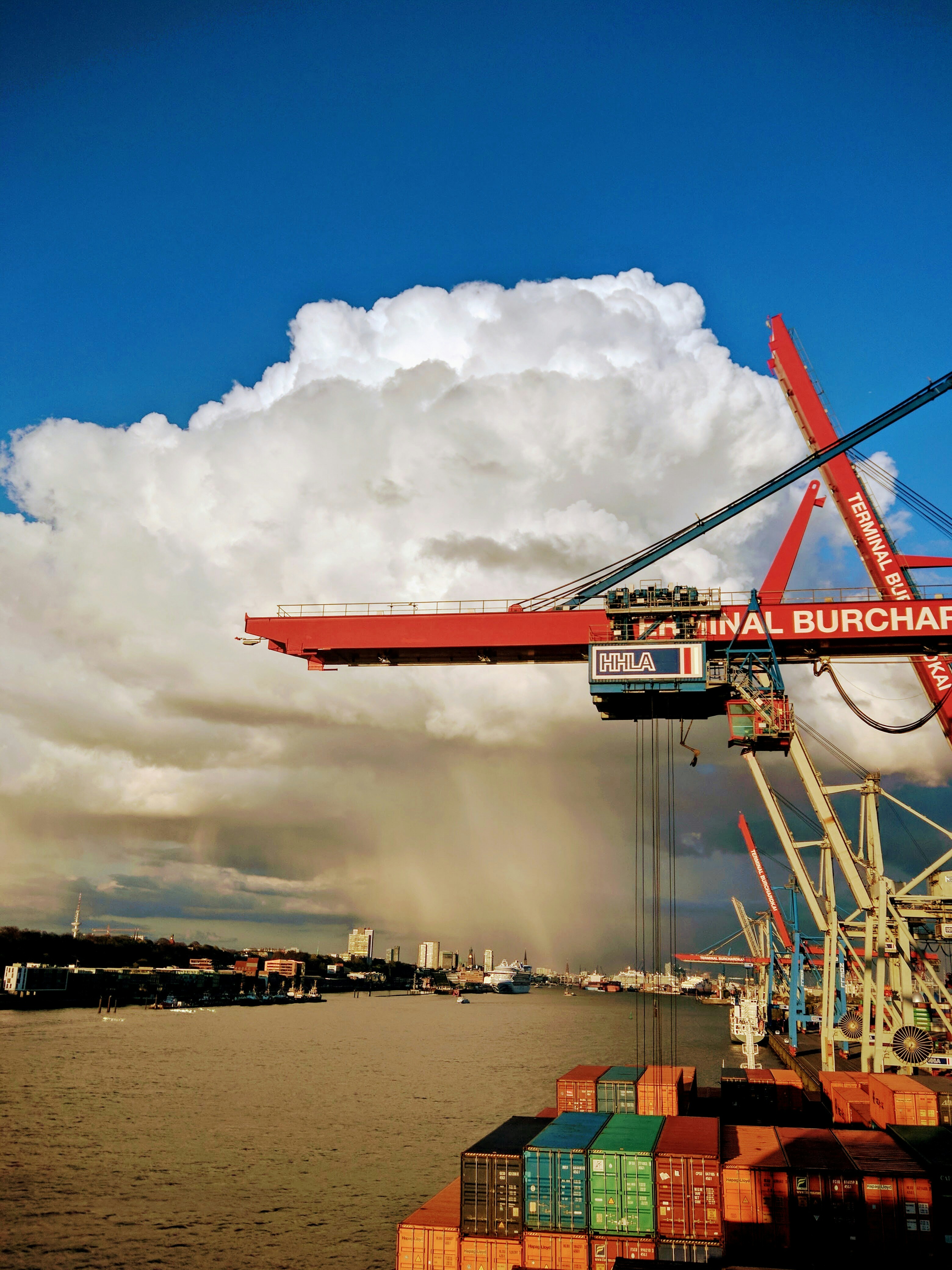 Container ship docked at a port with towering cranes against a backdrop of dramatic clouds and a distant city skyline.