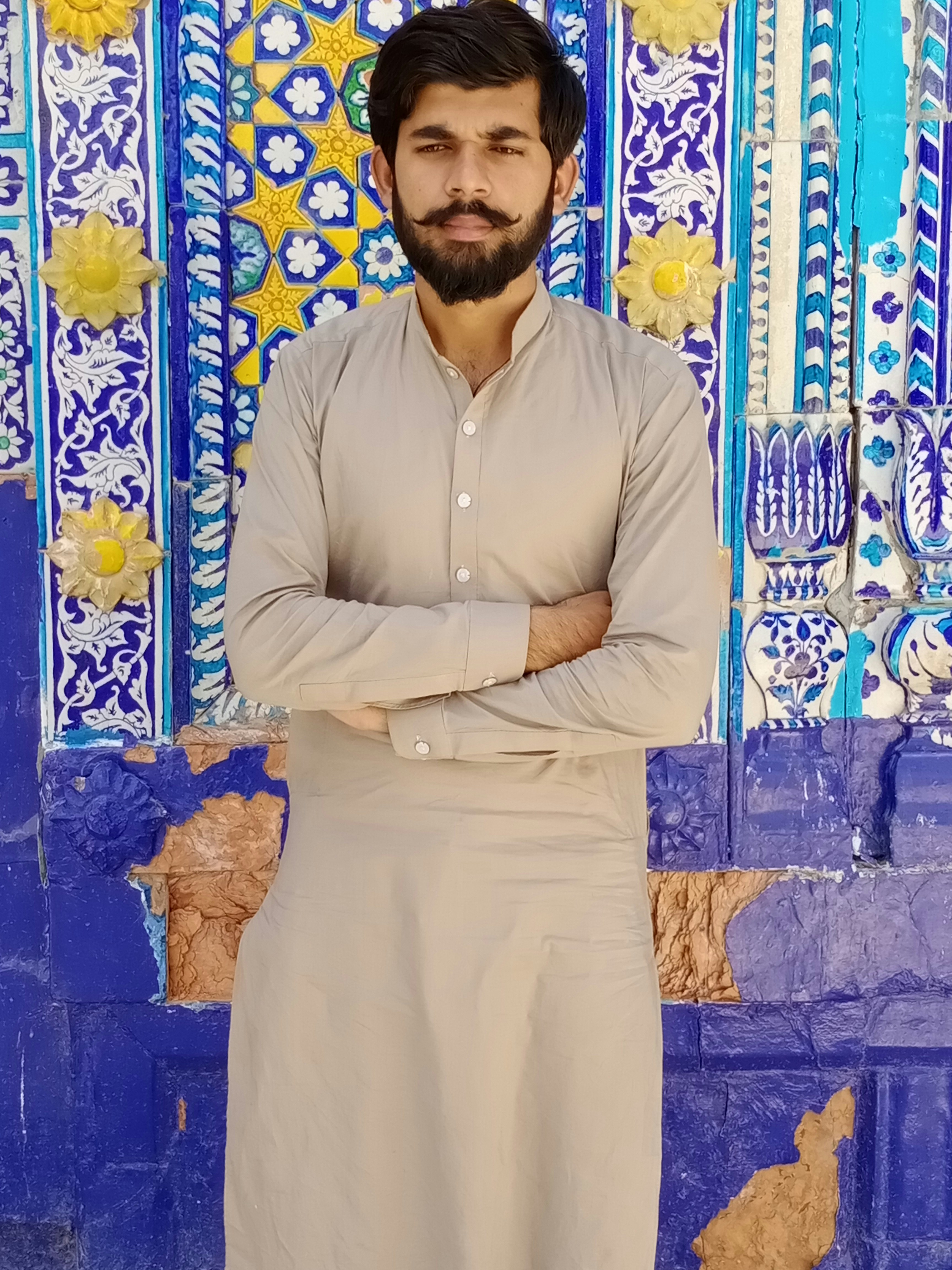 Man in traditional attire stands confidently against a richly decorated wall featuring intricate blue tile work and floral motifs.