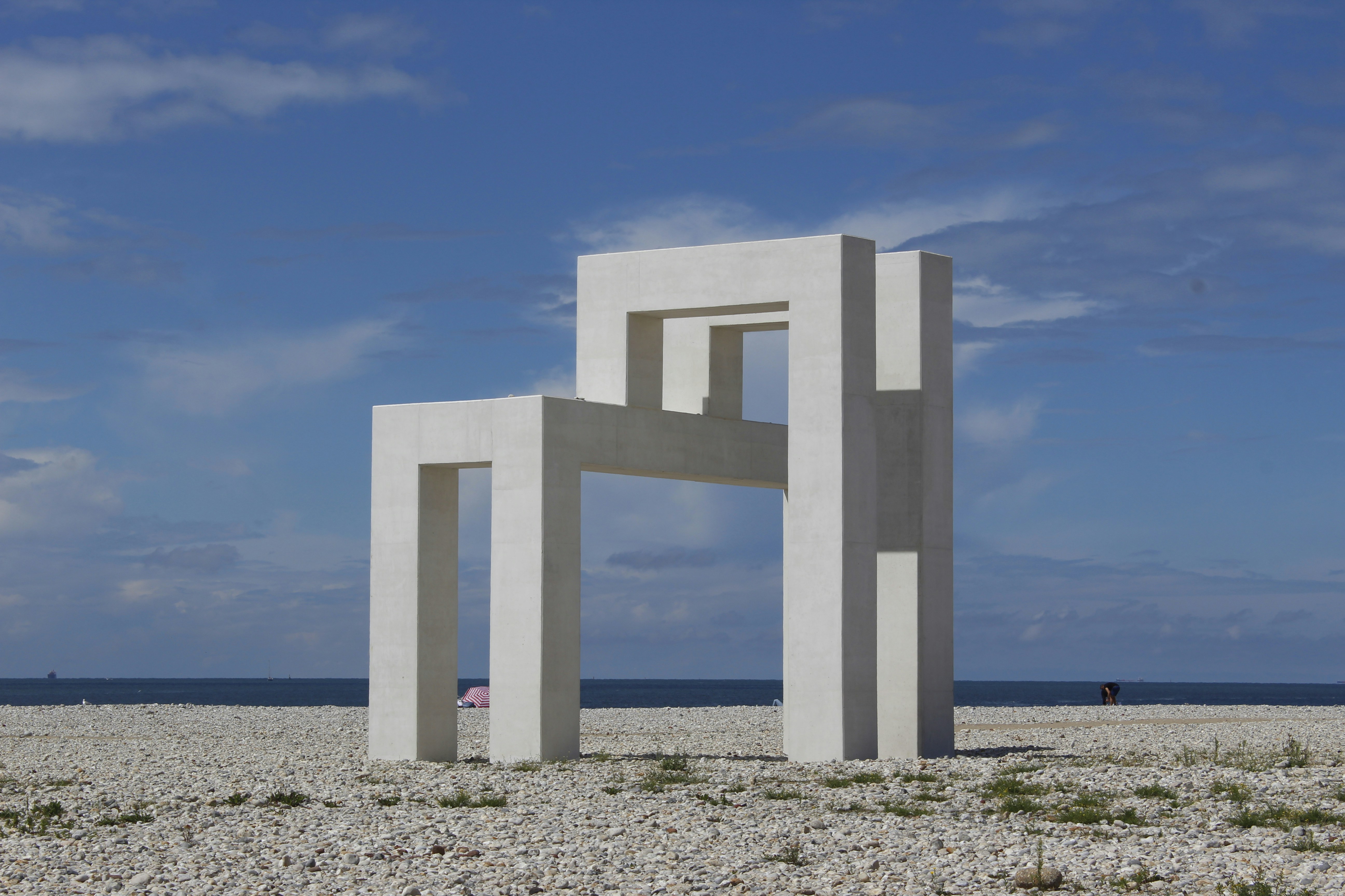 Large white geometric sculpture on a rocky beach under a bright blue sky.