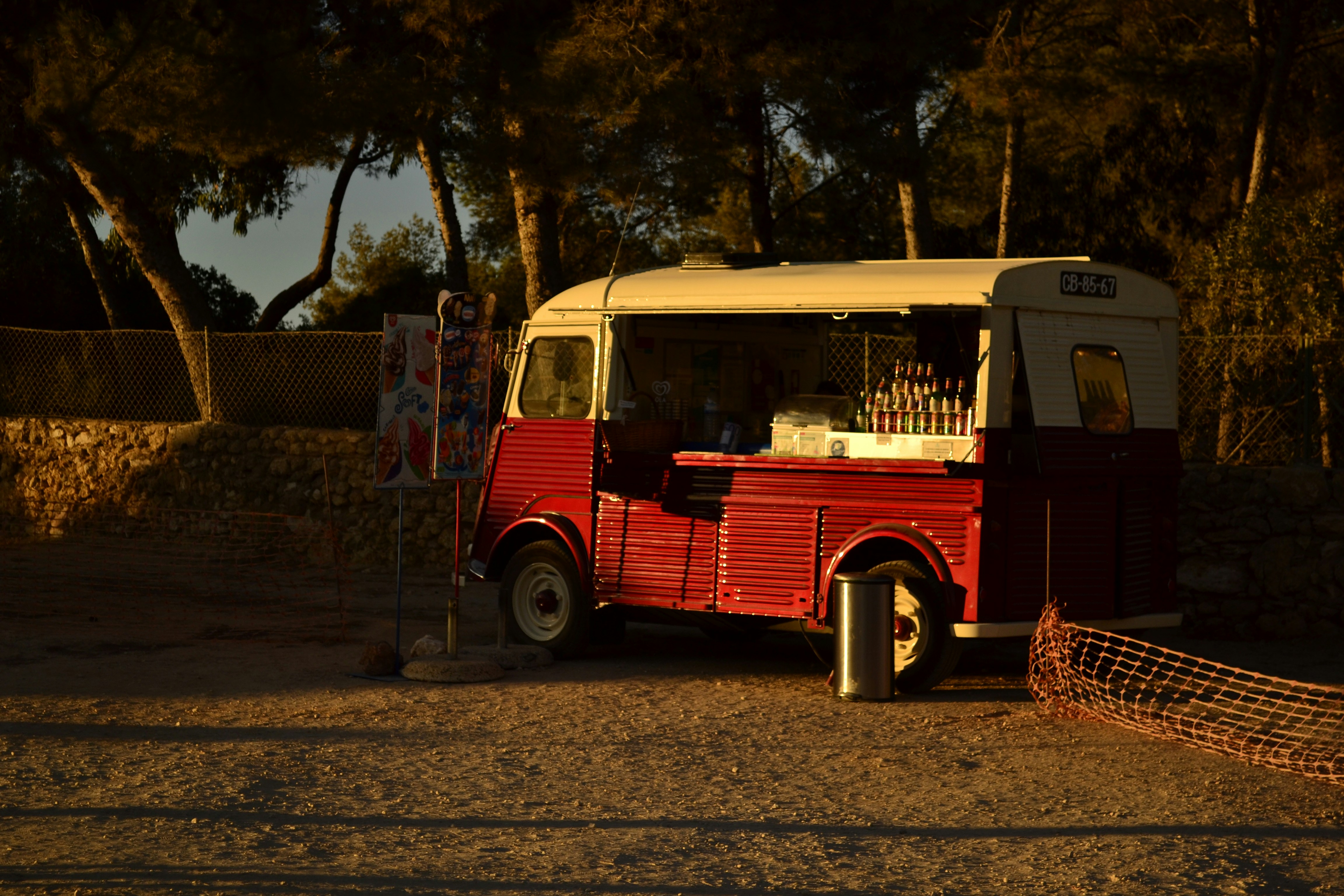 Charming retro food truck parked under the trees, basking in the warm glow of sunset. A vibrant sign and assortment of drinks are visible.