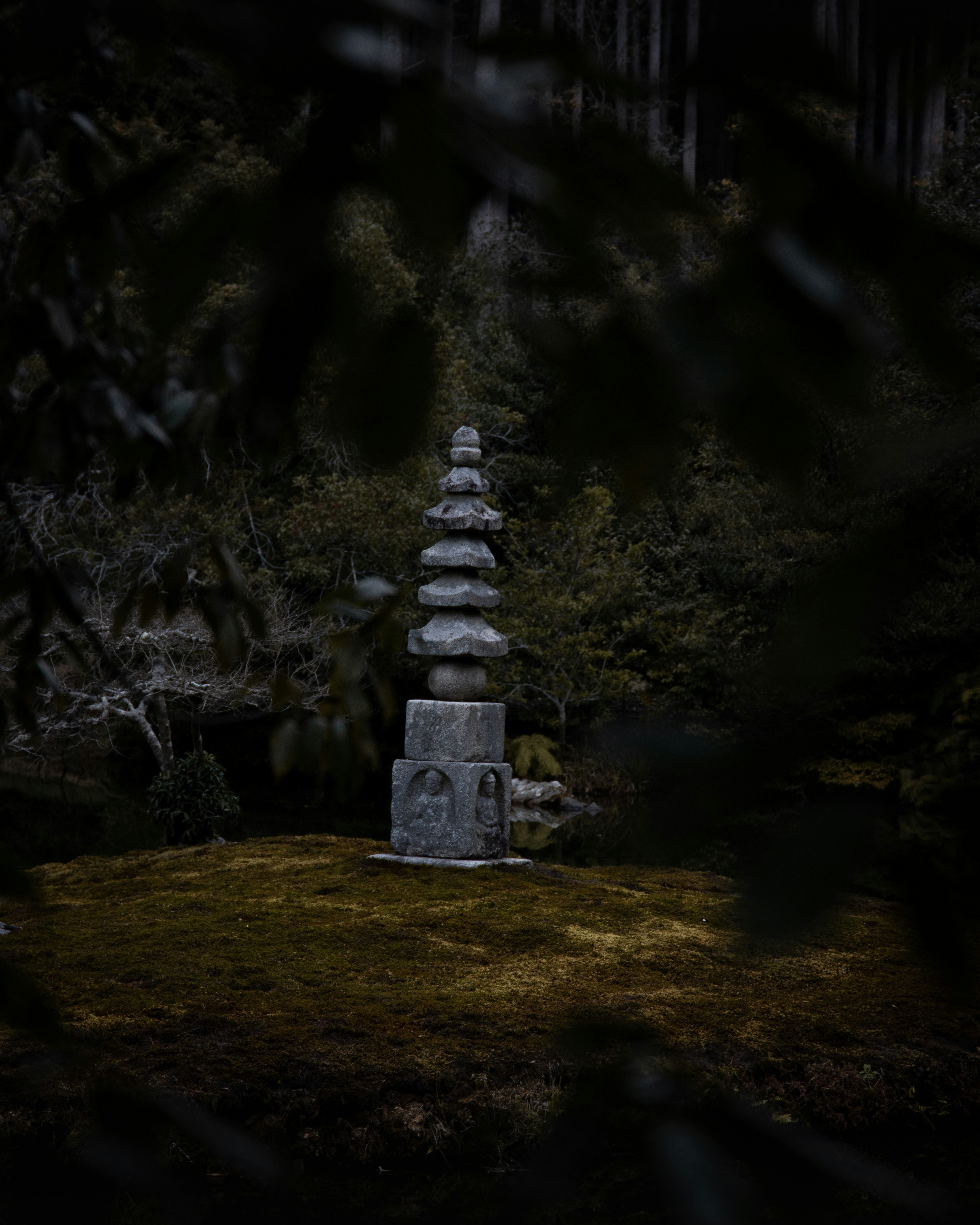 A serene stone pagoda stands amidst lush greenery, partially obscured by leaves, creating a sense of mystery and calm. The tranquil setting invites contemplation.
