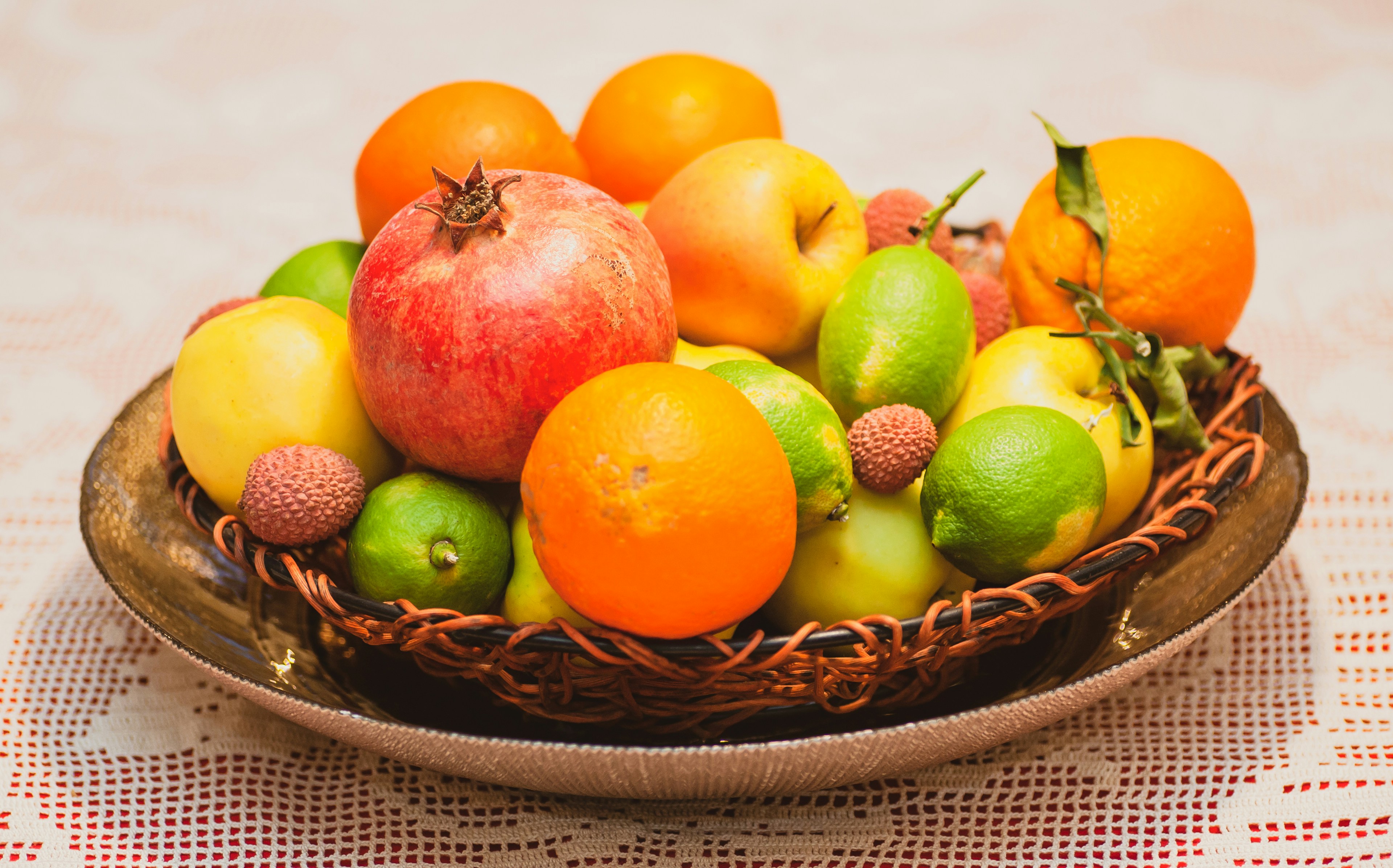 A woven basket brimming with a variety of fruits, including pomegranates, oranges, and lychees, arranged on a delicate lace tablecloth.
