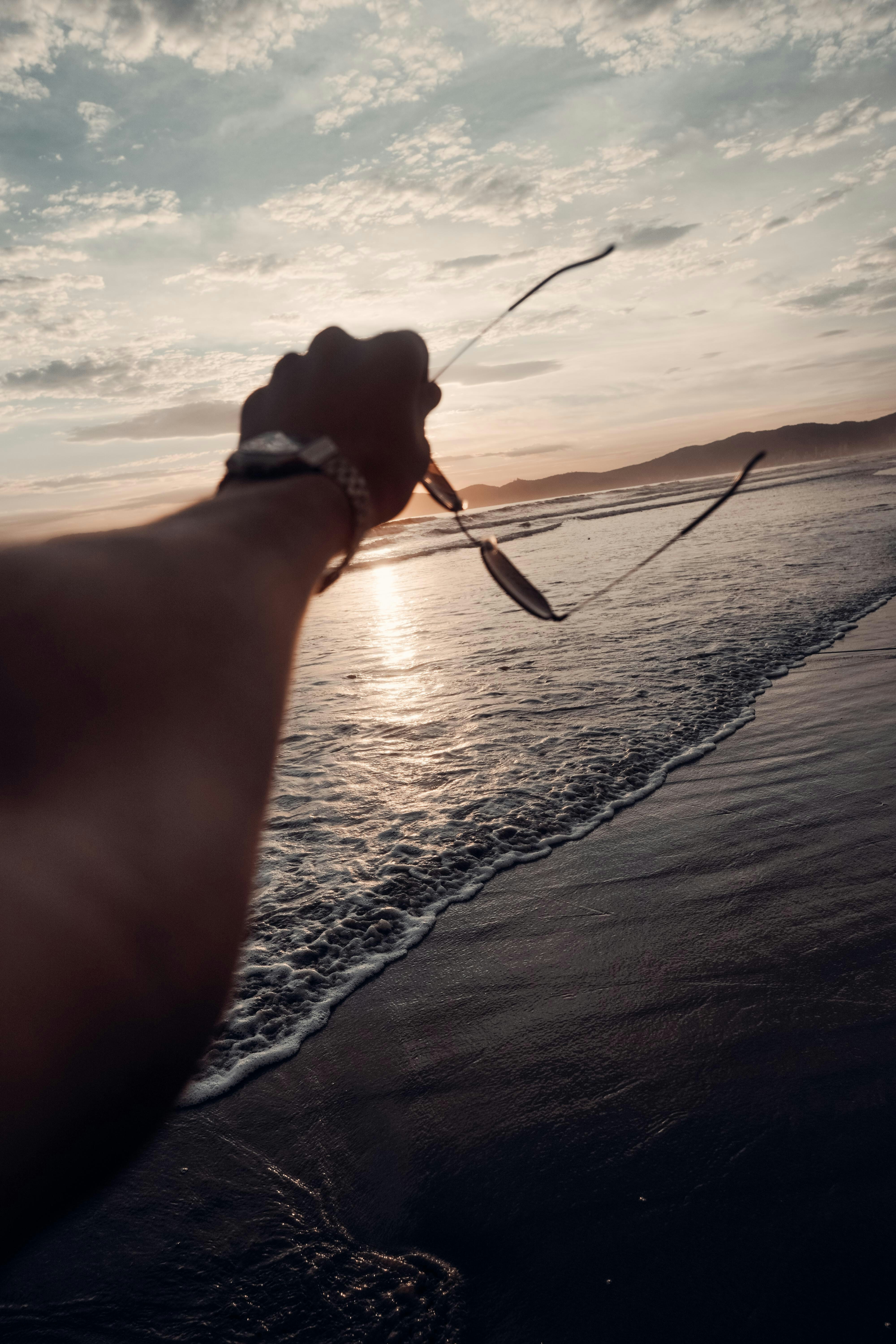 Chasing the Sunset: A Hand Reaches for the Shoreline with Sunglasses in Flight