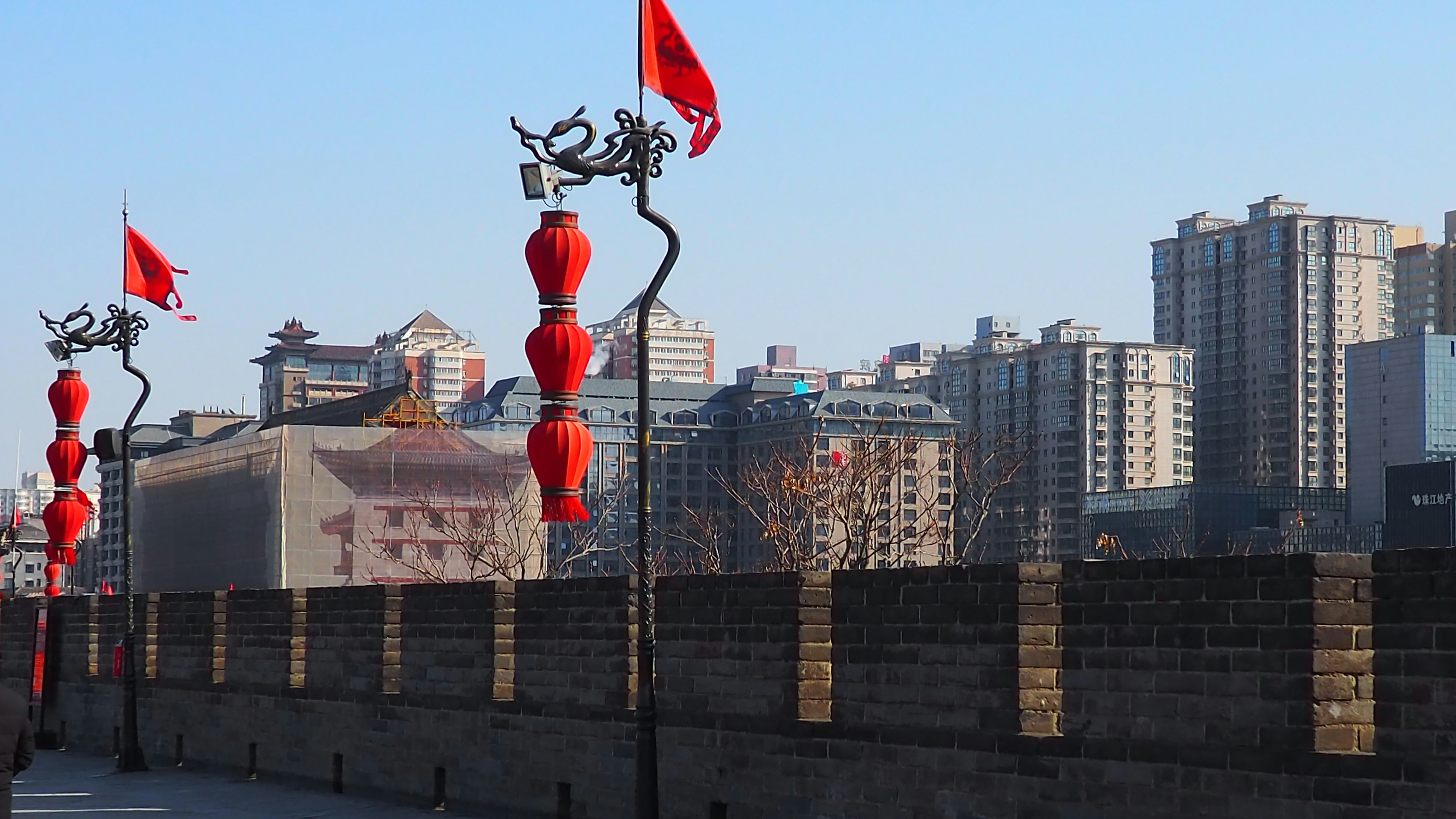 Traditional lanterns and vibrant red flags adorn a historic wall, juxtaposed against a backdrop of contemporary high-rise buildings.