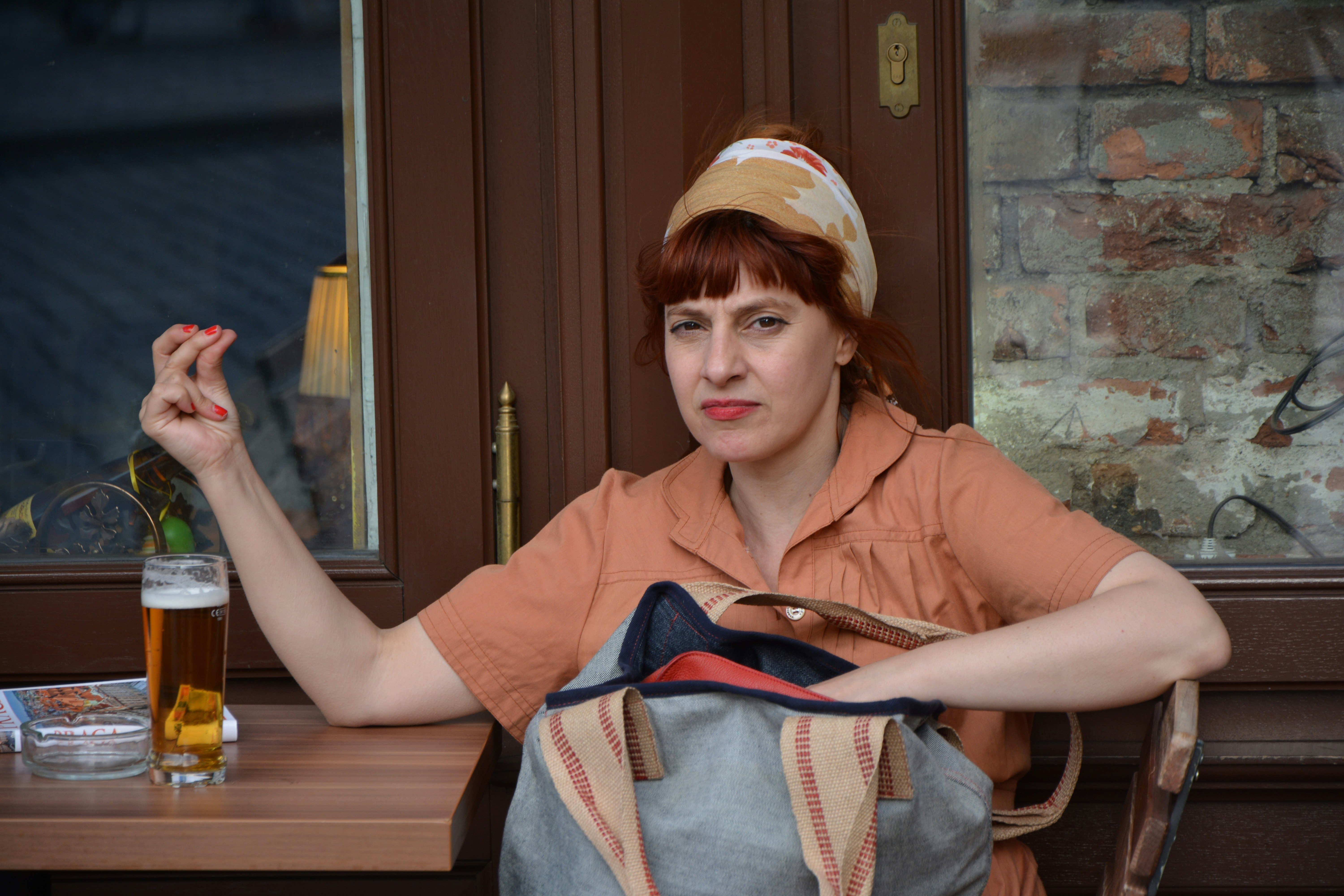 Woman sitting at a café table with a beer, expressing frustration while holding a handbag. The scene captures a candid moment in a bustling urban setting.