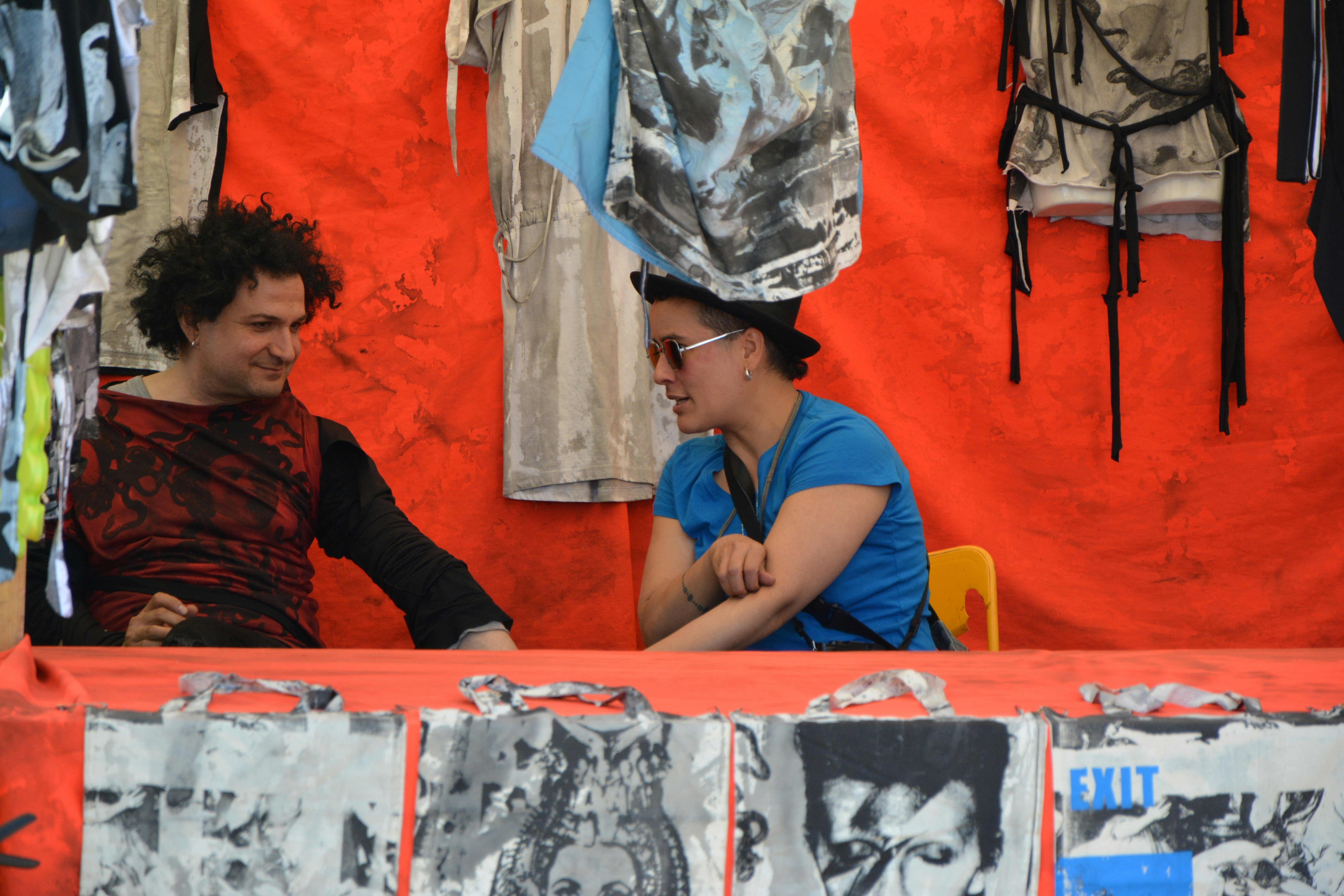 Two individuals engaged in conversation at a vibrant market stall adorned with artistic prints and clothing.
