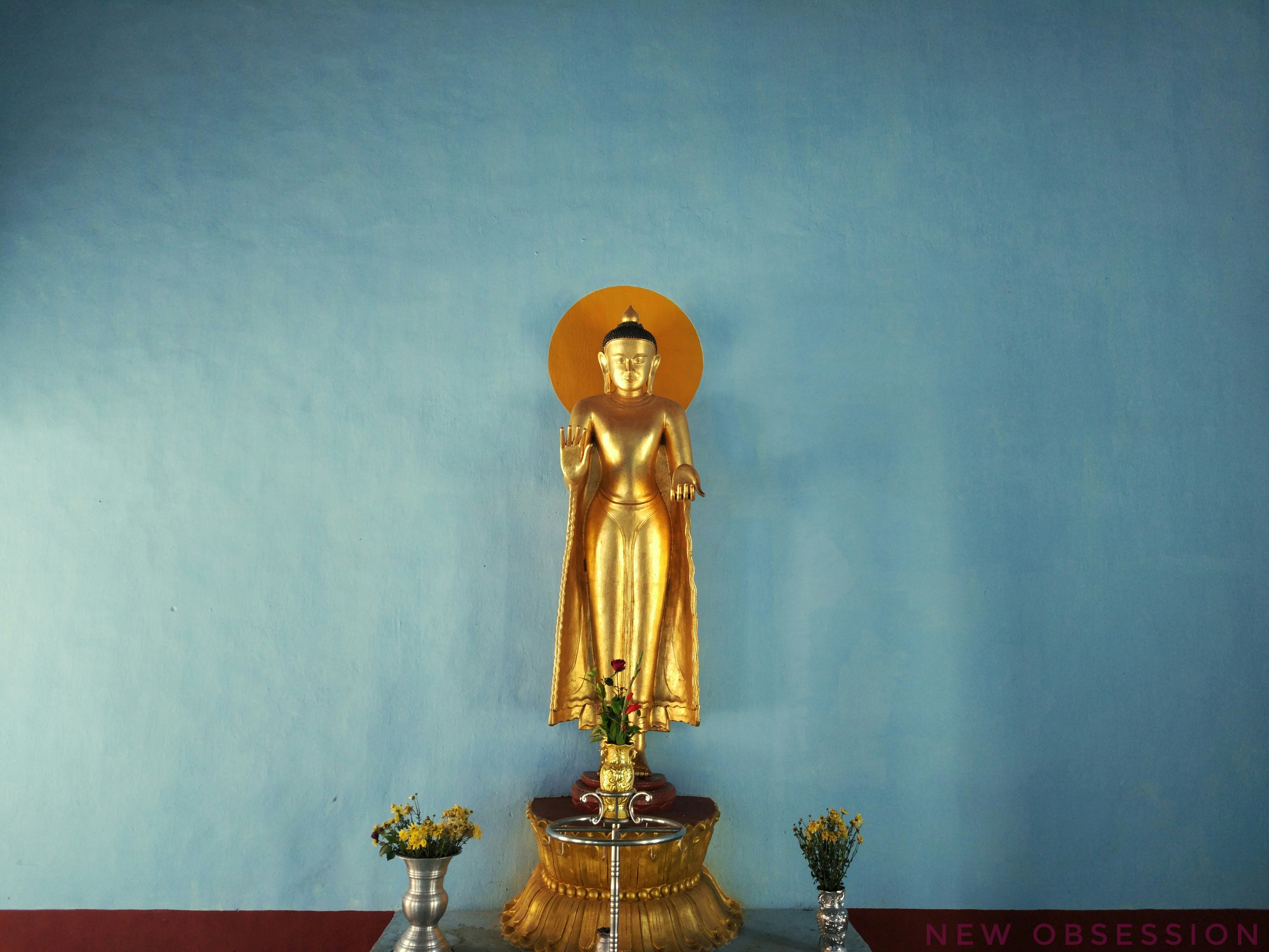 A golden Buddha statue stands on a raised pedestal against a blue wall, flanked by small flower arrangements.