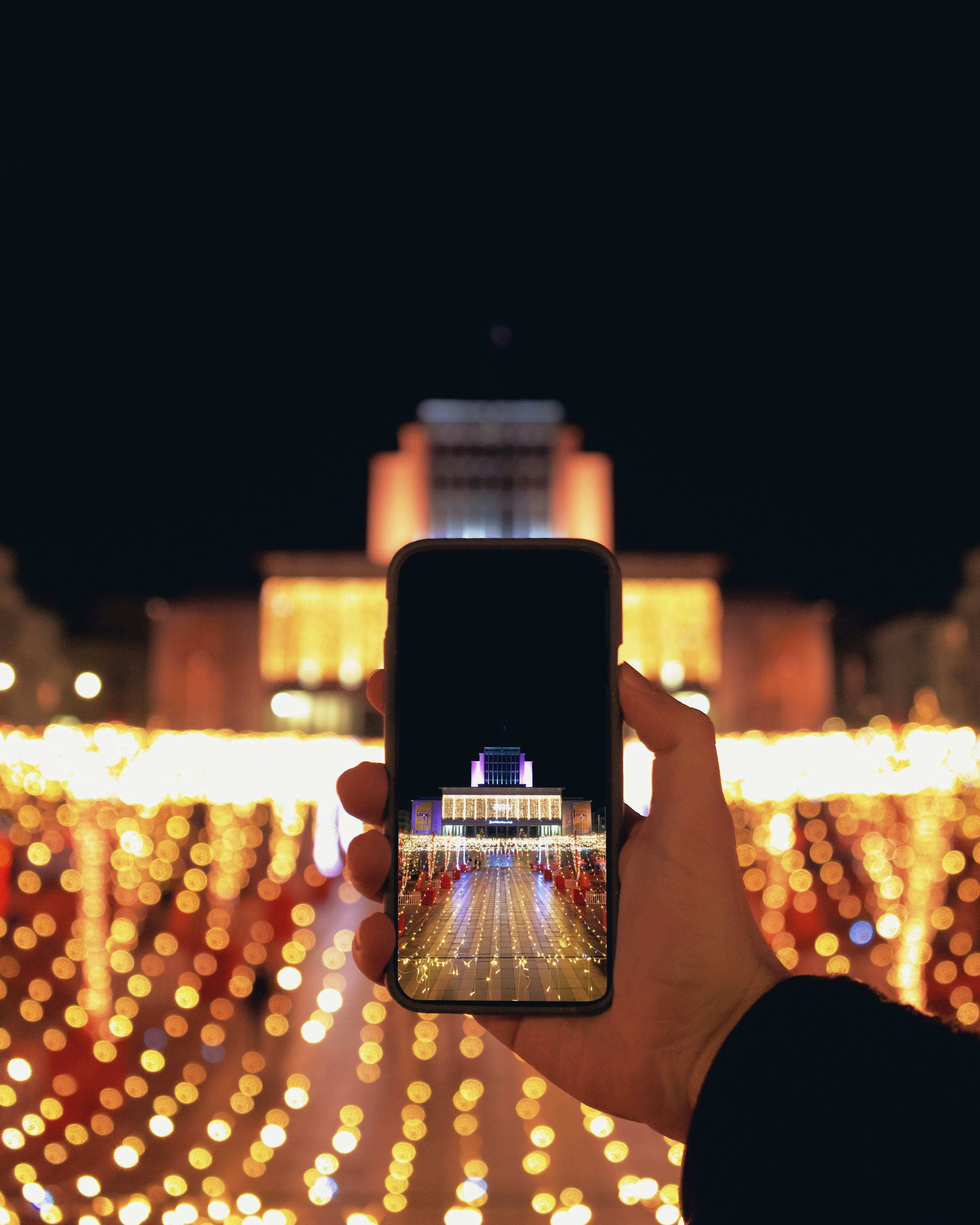 A hand holds a smartphone displaying a brightly lit building against a backdrop of festive lights. The scene captures the contrast between the digital and physical worlds.