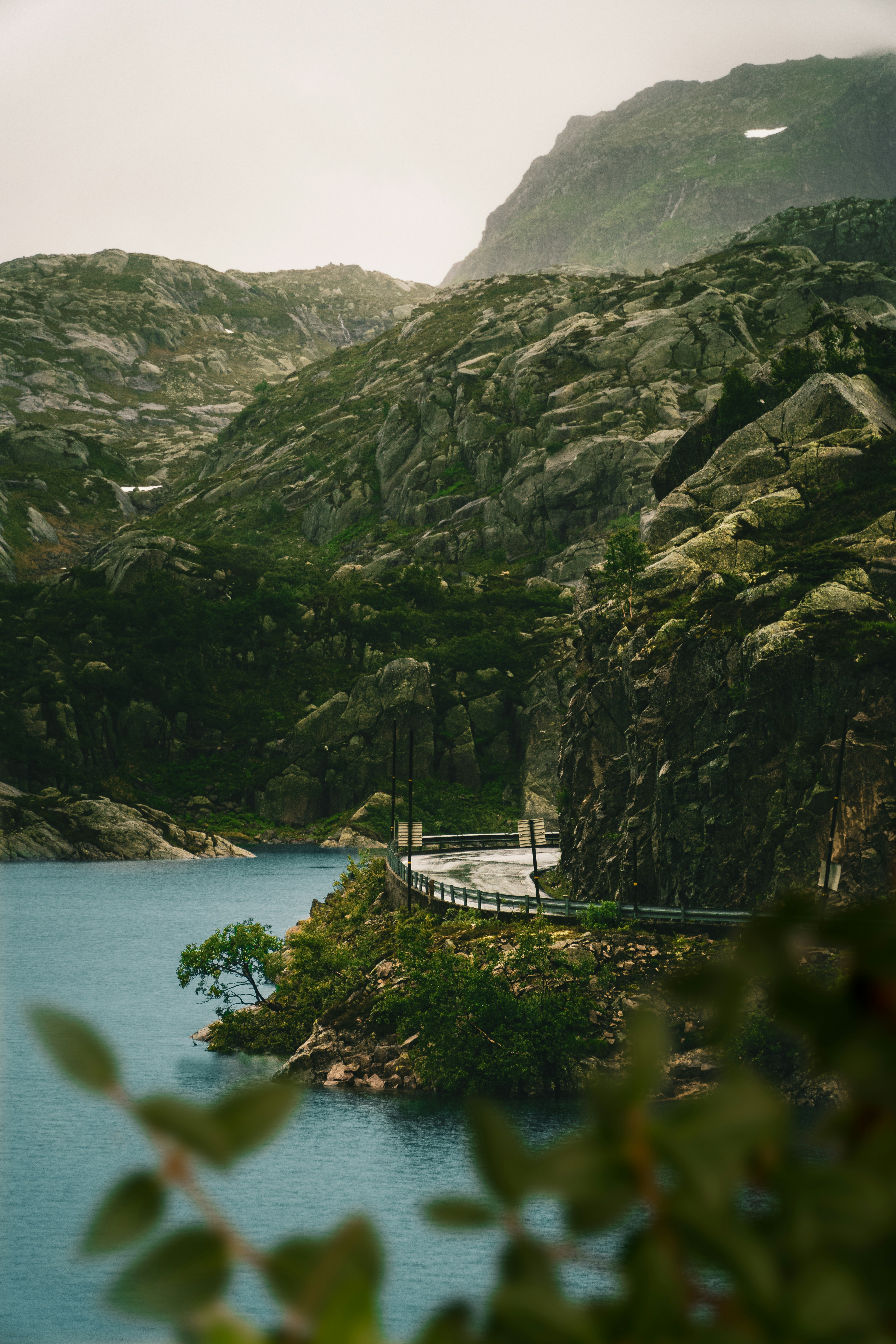 Serpentine road weaving through lush, rocky hills beside a tranquil lake under a cloudy sky.