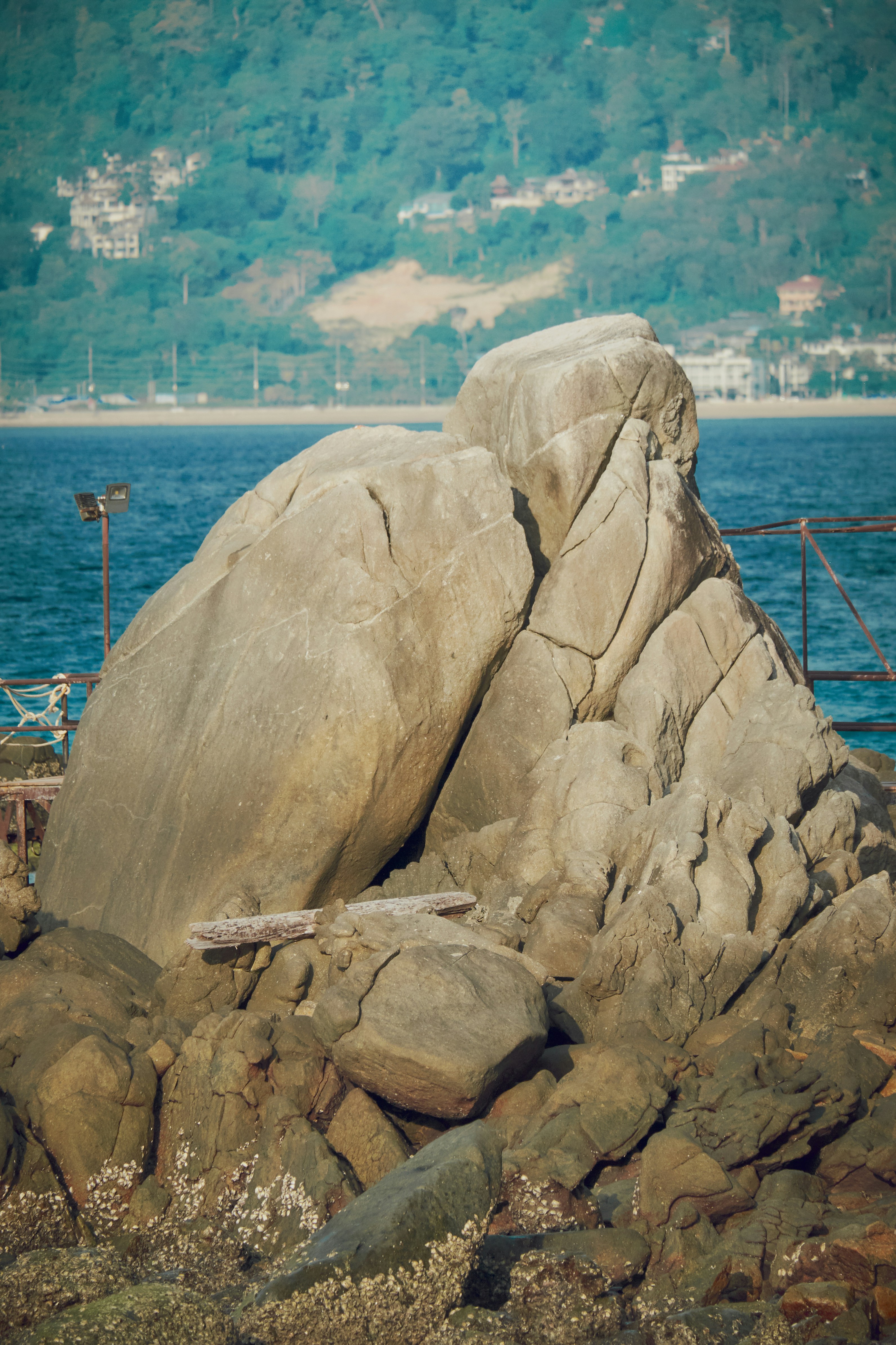 Large rock formations stand resolutely at the water's edge, with a serene ocean backdrop and distant hills visible. A wooden plank rests on one of the rocks.