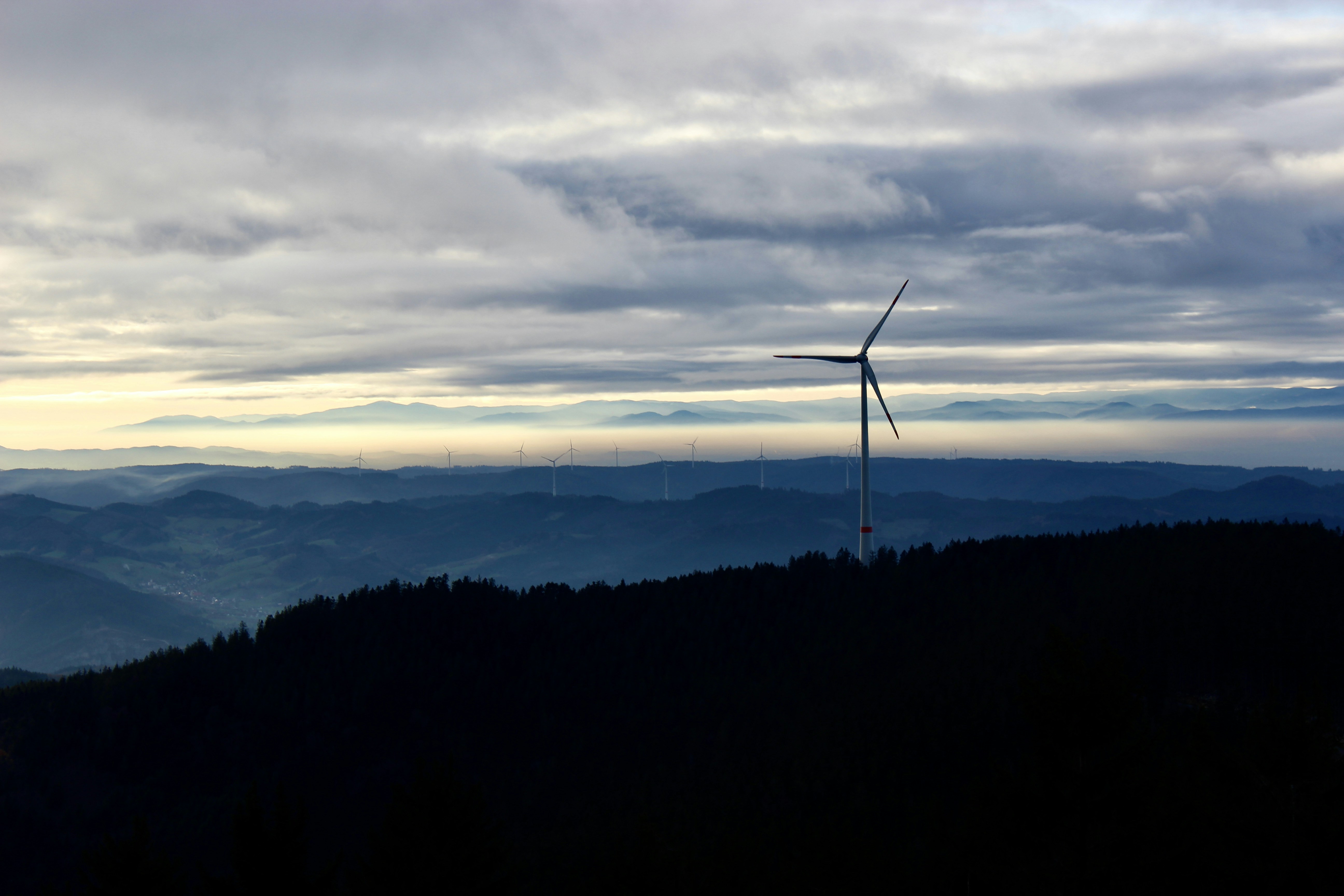 Wind turbine silhouetted against layered mountain ranges under a cloud-laden sky at dawn.