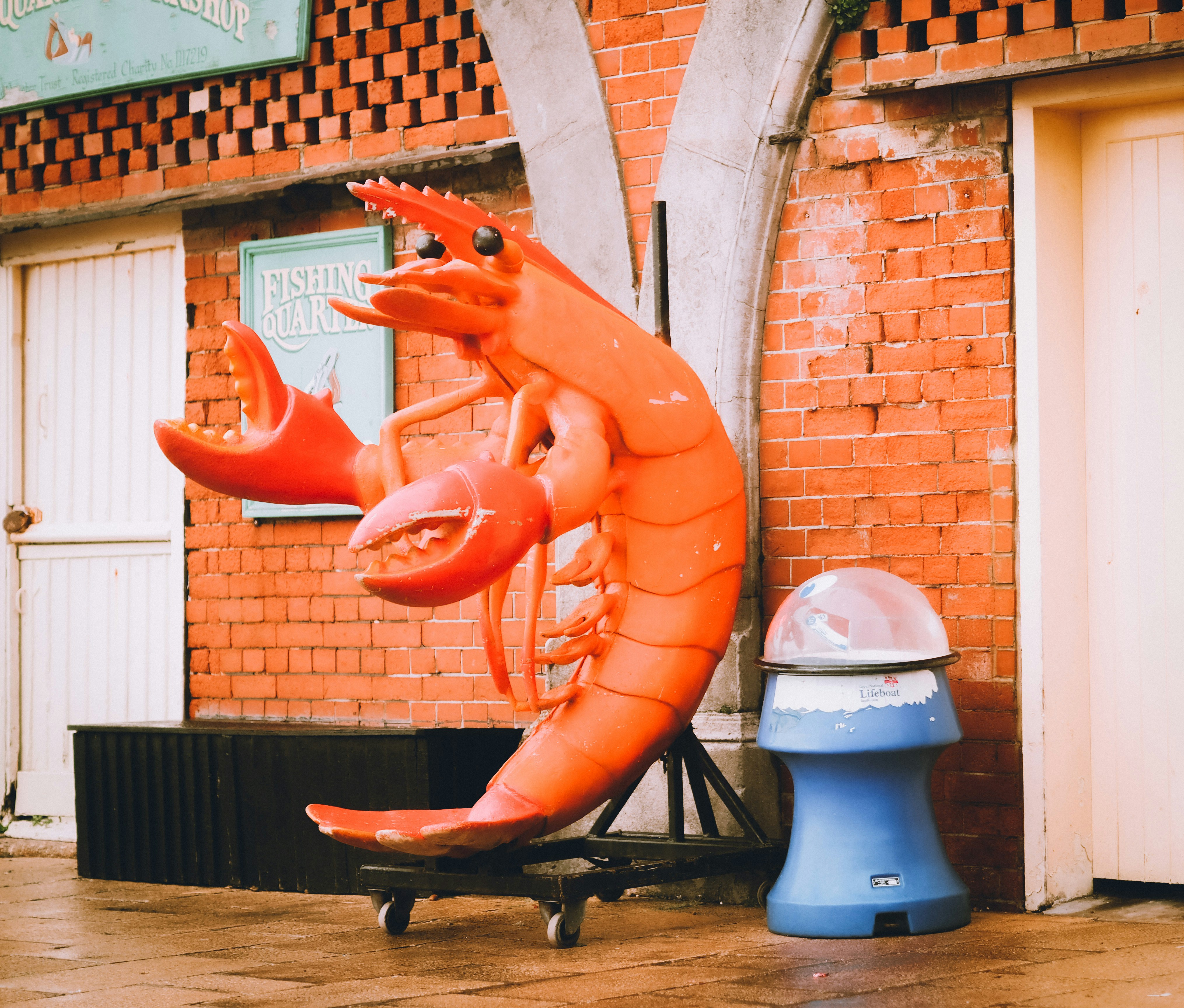 A vibrant, oversized lobster sculpture positioned against a brick wall, showcasing its claws prominently beside a whimsical trash can.