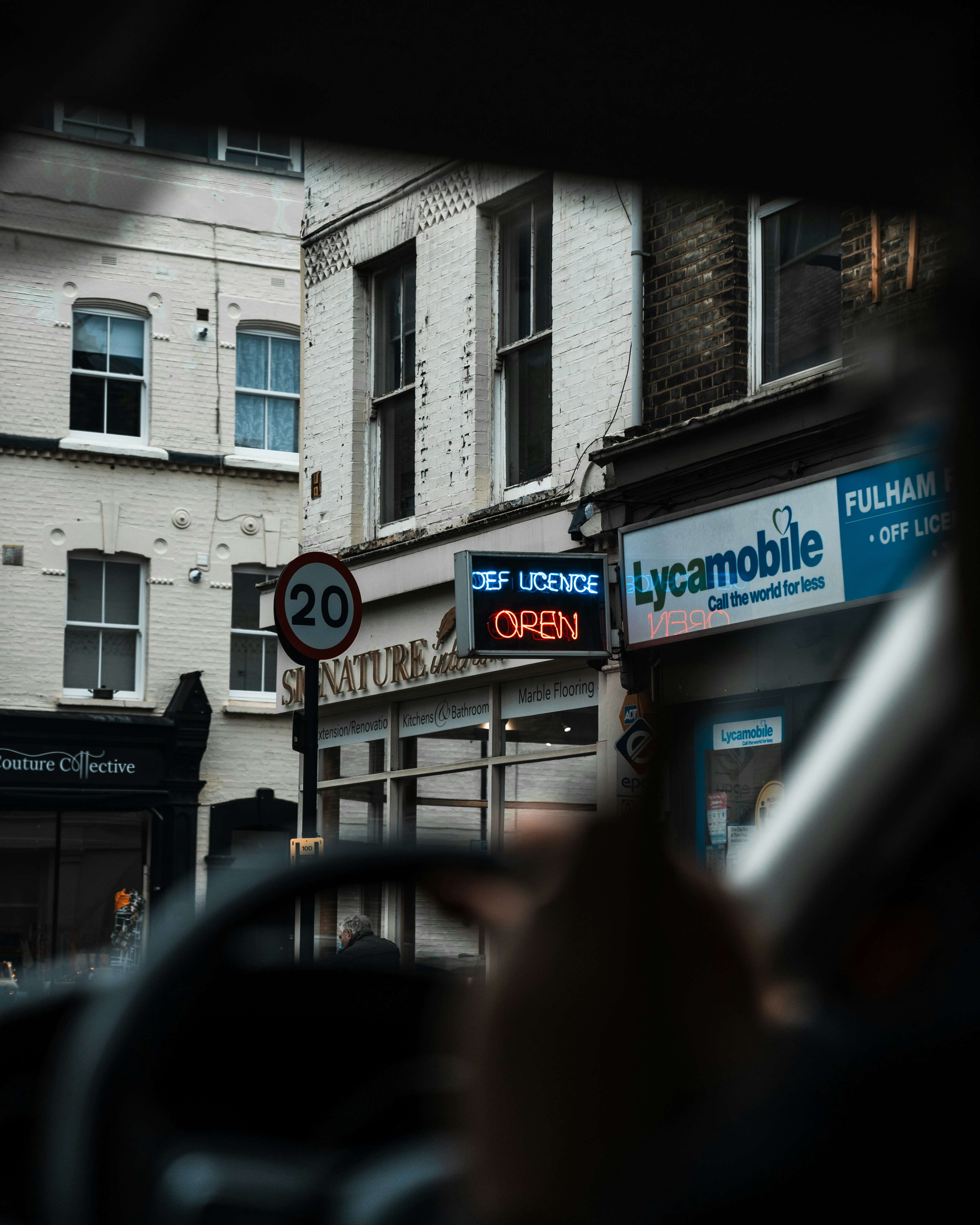 A vibrant off-licence shop sign illuminated in neon, framed by urban architecture, capturing a slice of city life. The scene is viewed from inside a vehicle, adding context to the bustling environment.