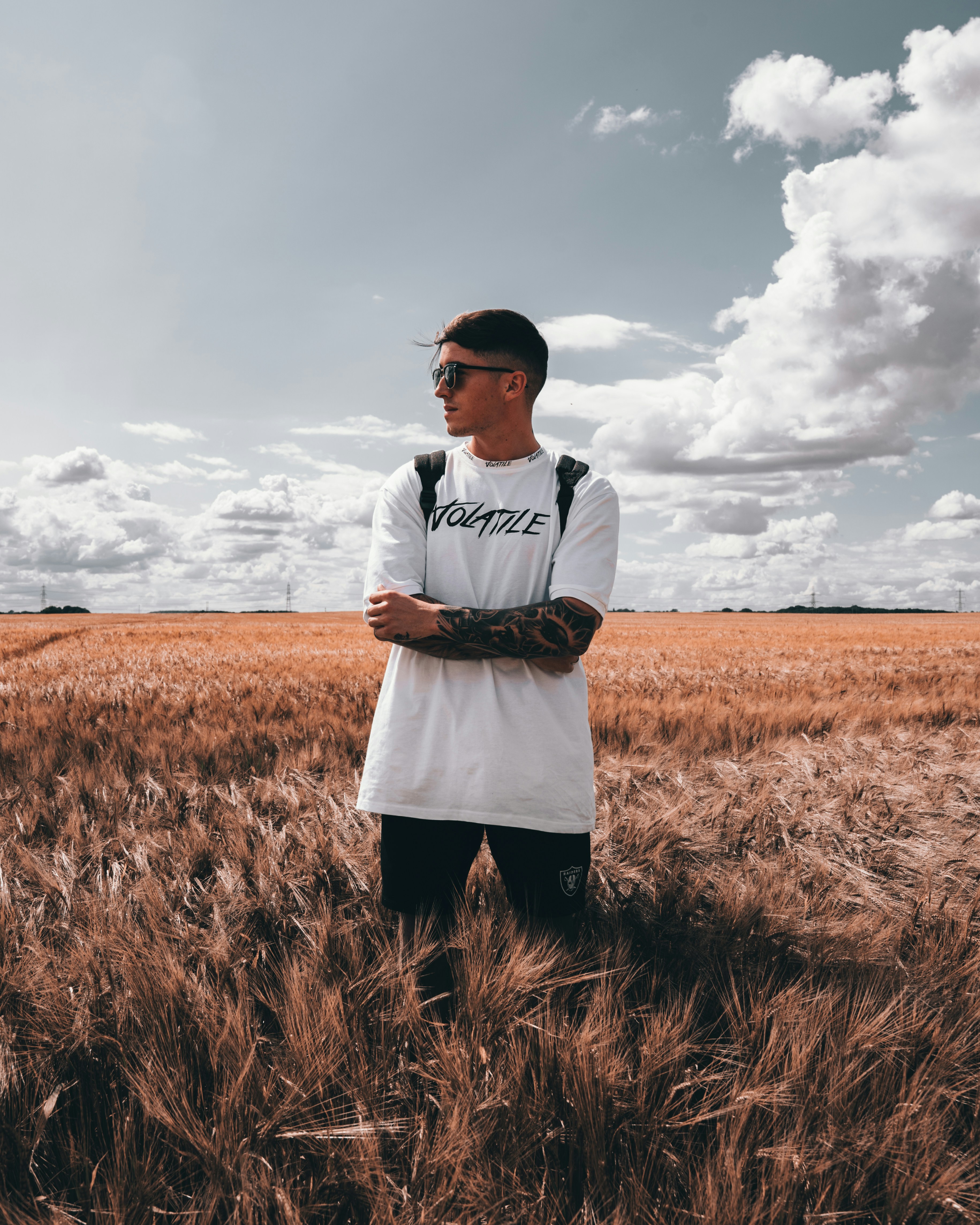 Young man standing confidently in a golden wheat field under a dramatic sky, wearing a white t-shirt with bold lettering.