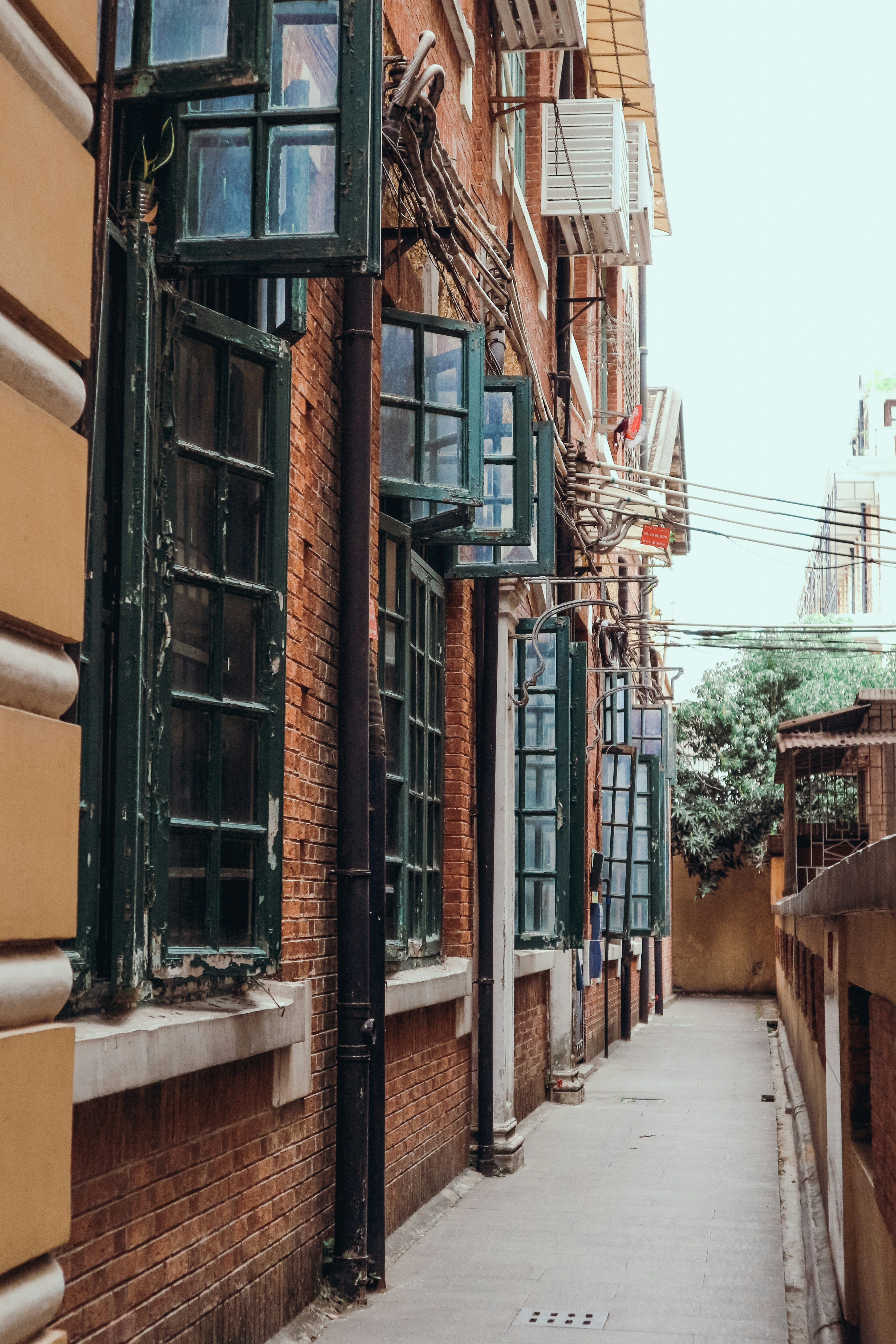 Charming alleyway flanked by vintage brick buildings with open green windows, revealing glimpses of urban life. A serene path leads through the historical architecture.