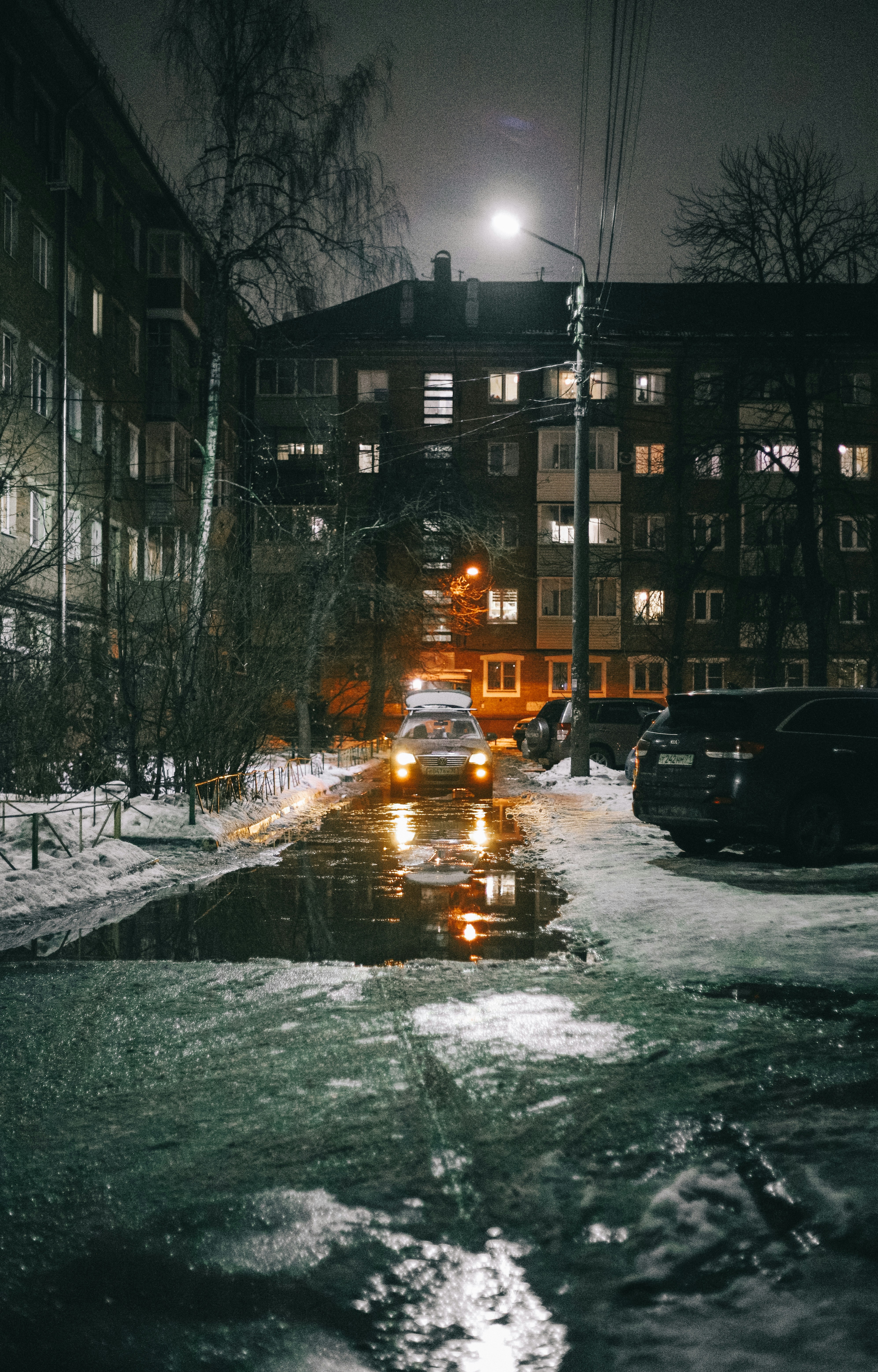 A car navigates a flooded street illuminated by streetlights, with reflections shimmering on the water's surface. The surrounding buildings loom in the background.