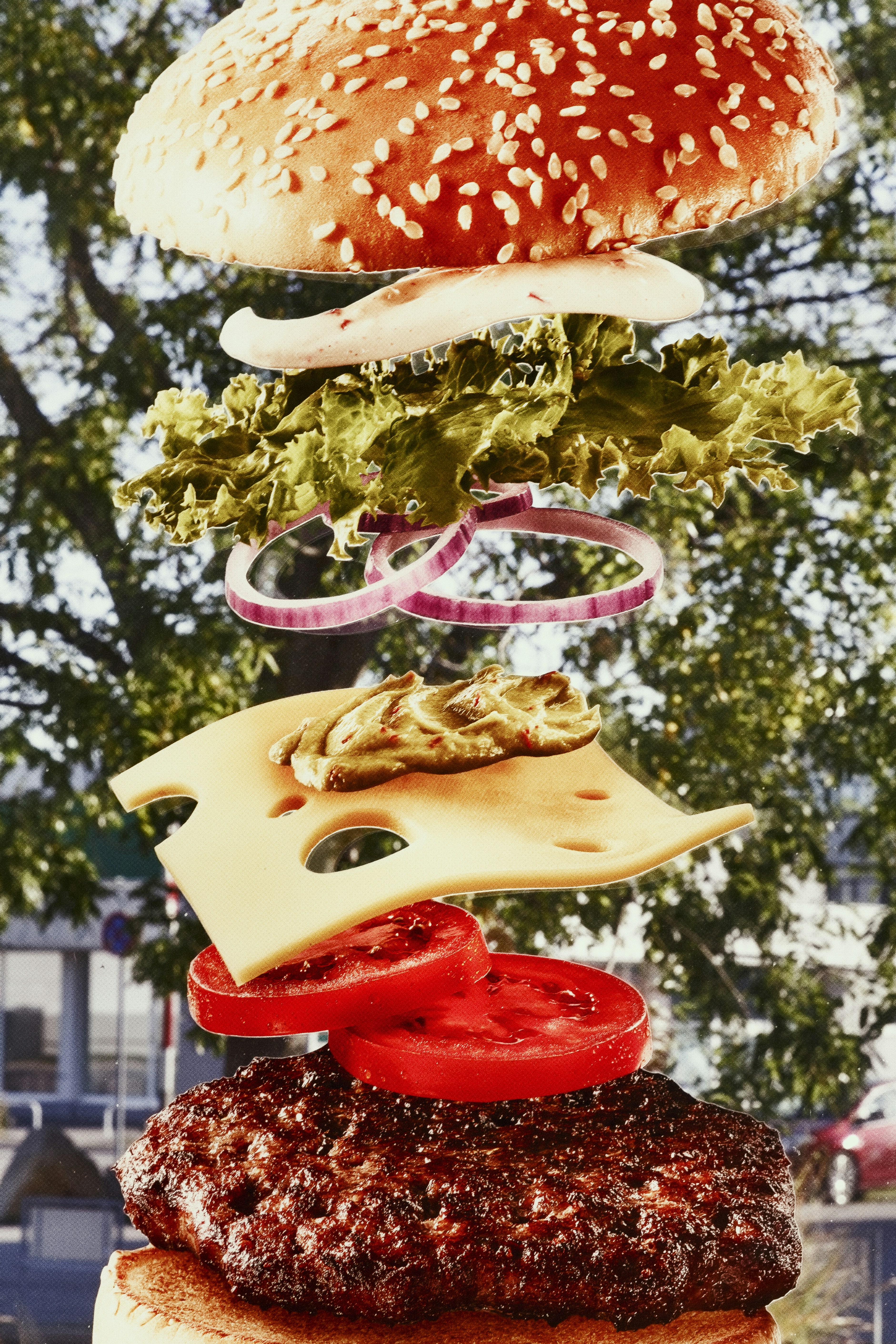 Floating layers of a hamburger, showcasing fresh ingredients like lettuce, tomato, cheese, and pickles against a blurred outdoor backdrop.