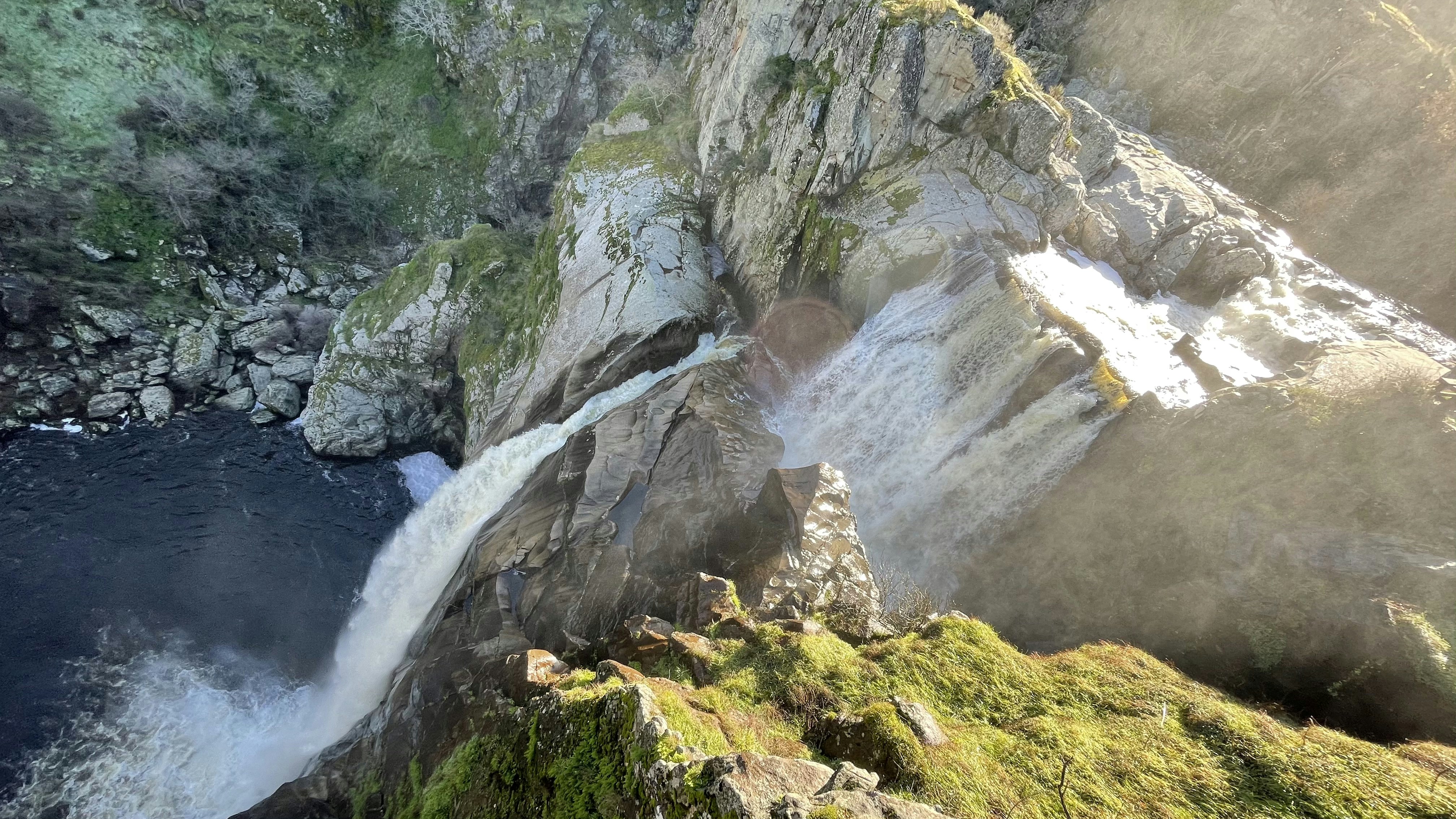 Waterfall plunging into a rocky pool surrounded by lush greenery under bright sunlight.