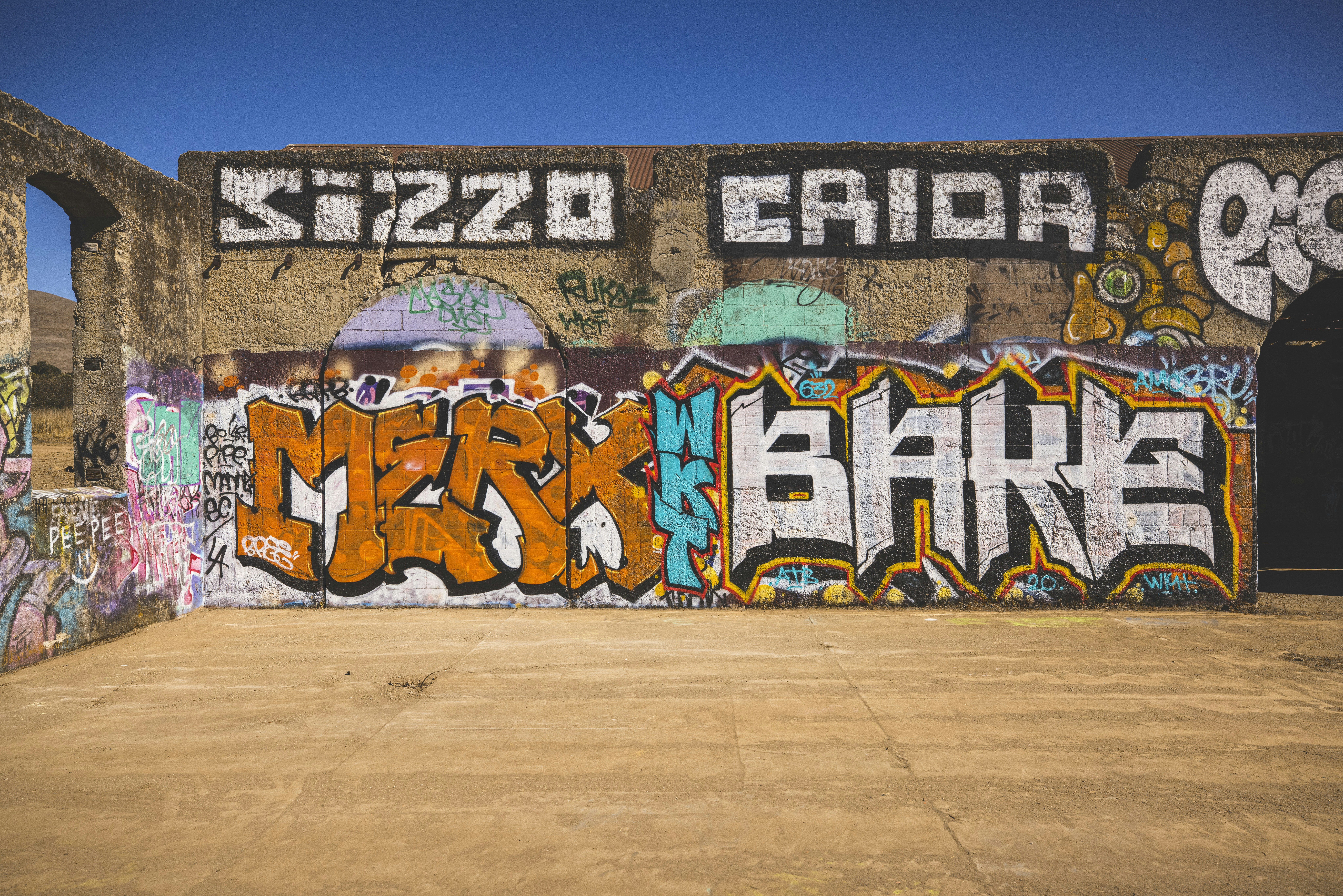 Colorful graffiti mural featuring the names 'MERK' and 'BAKE' on a weathered wall, surrounded by various tags and symbols. The artwork reflects the dynamic nature of urban expression.
