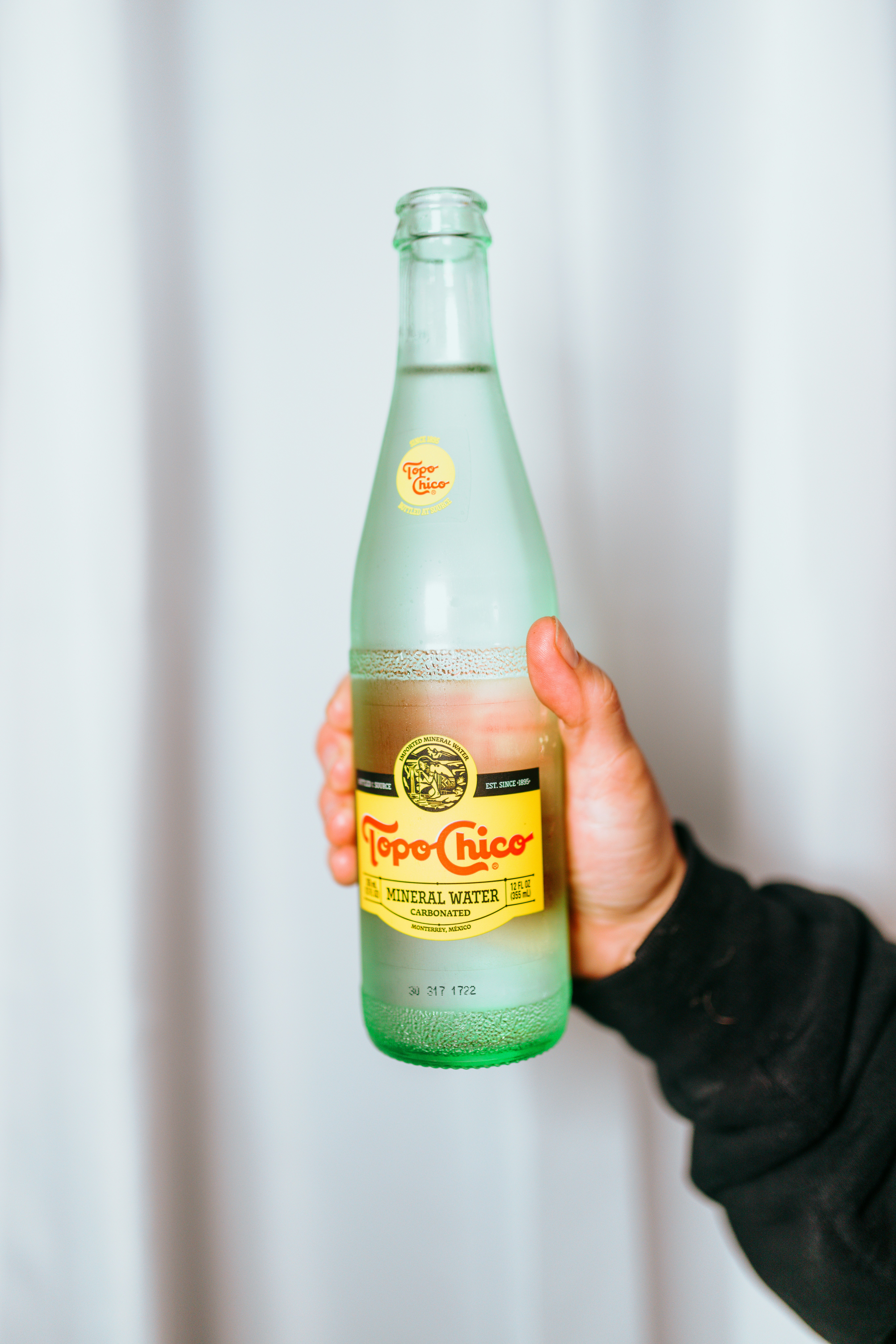 Hand holding a bottle of Topo Chico mineral water against a softly blurred background. The bottle showcases its distinctive label and refreshing contents.