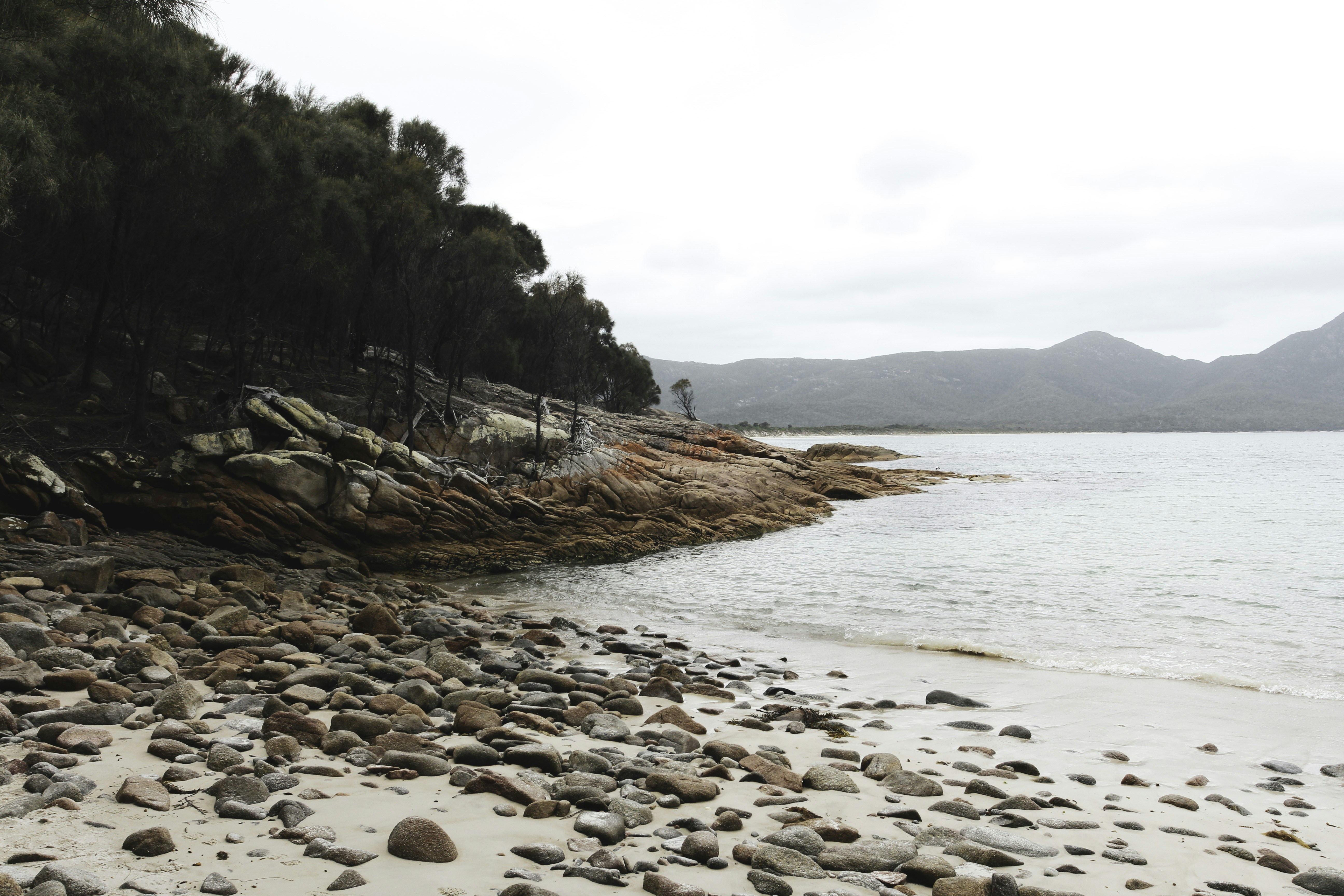 Rocky shoreline meeting calm waters under an overcast sky, with distant mountains visible.
