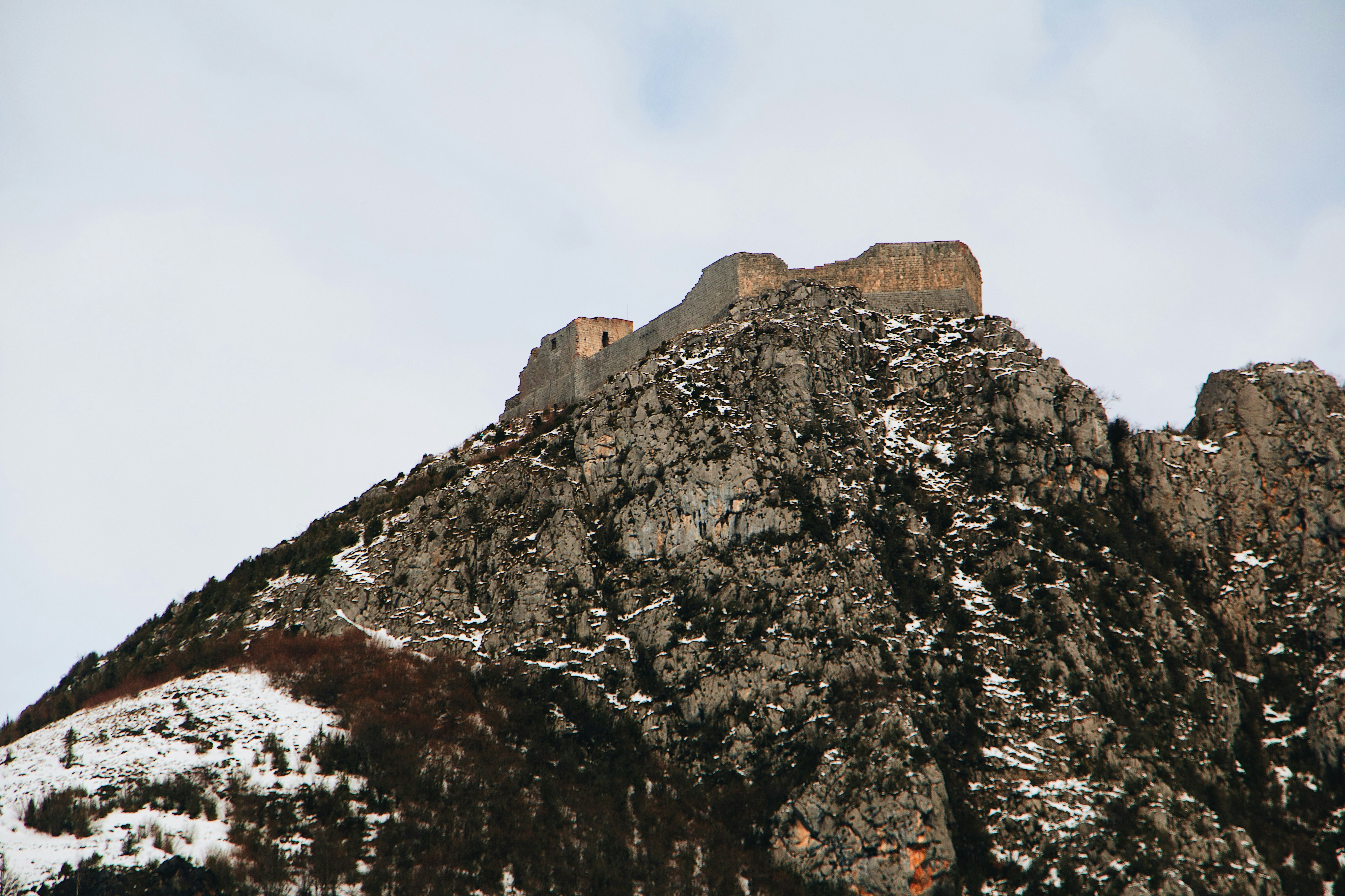 Ancient fortress perched atop a rocky mountain, surrounded by snow-capped terrain under a cloudy sky.