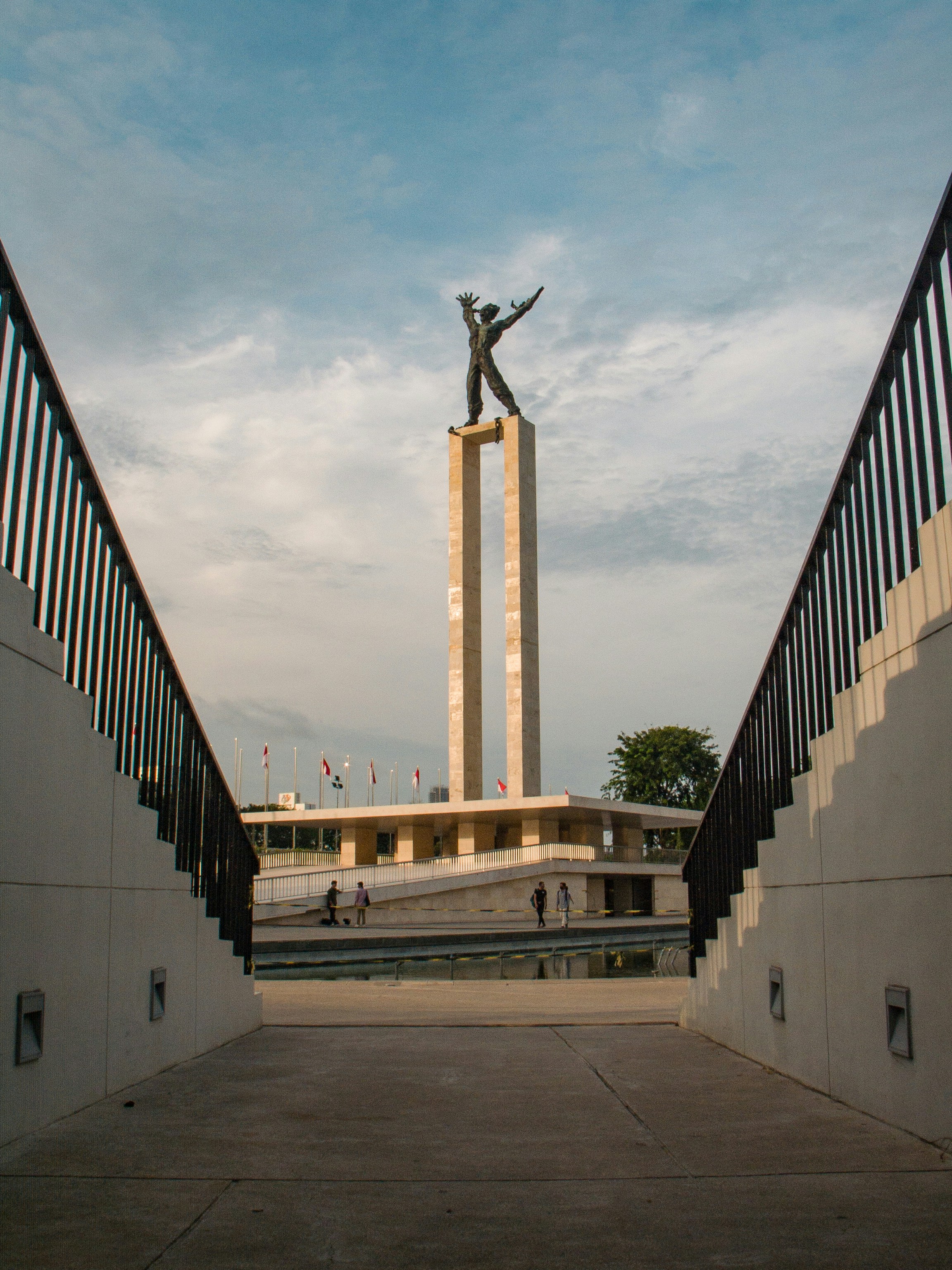 A towering statue of a figure reaching towards the sky, framed by stairways, symbolizing hope and aspiration. The setting features a serene backdrop with flags fluttering in the distance.