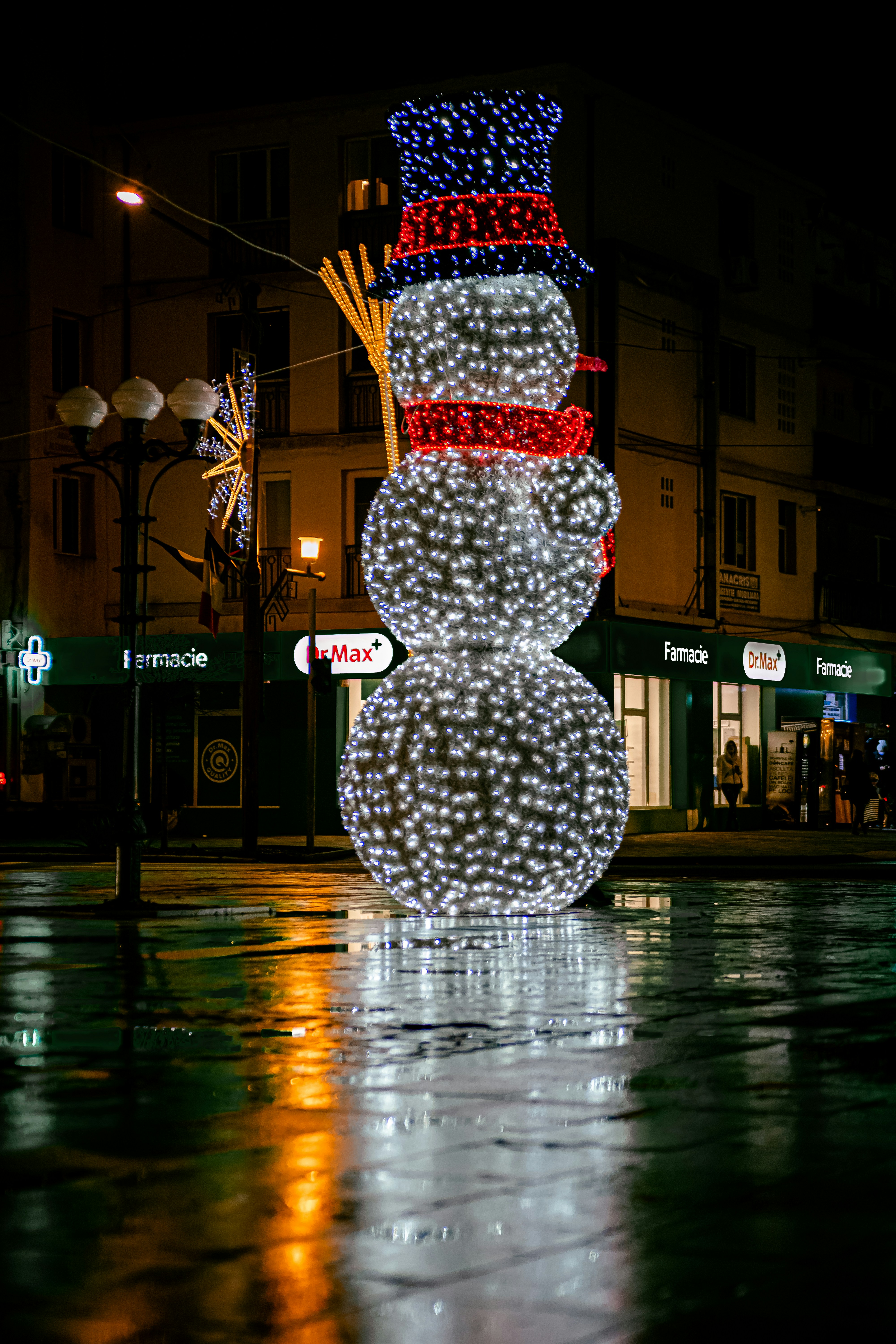 Brightly lit snowman decoration standing on a wet street, reflecting city lights at night.