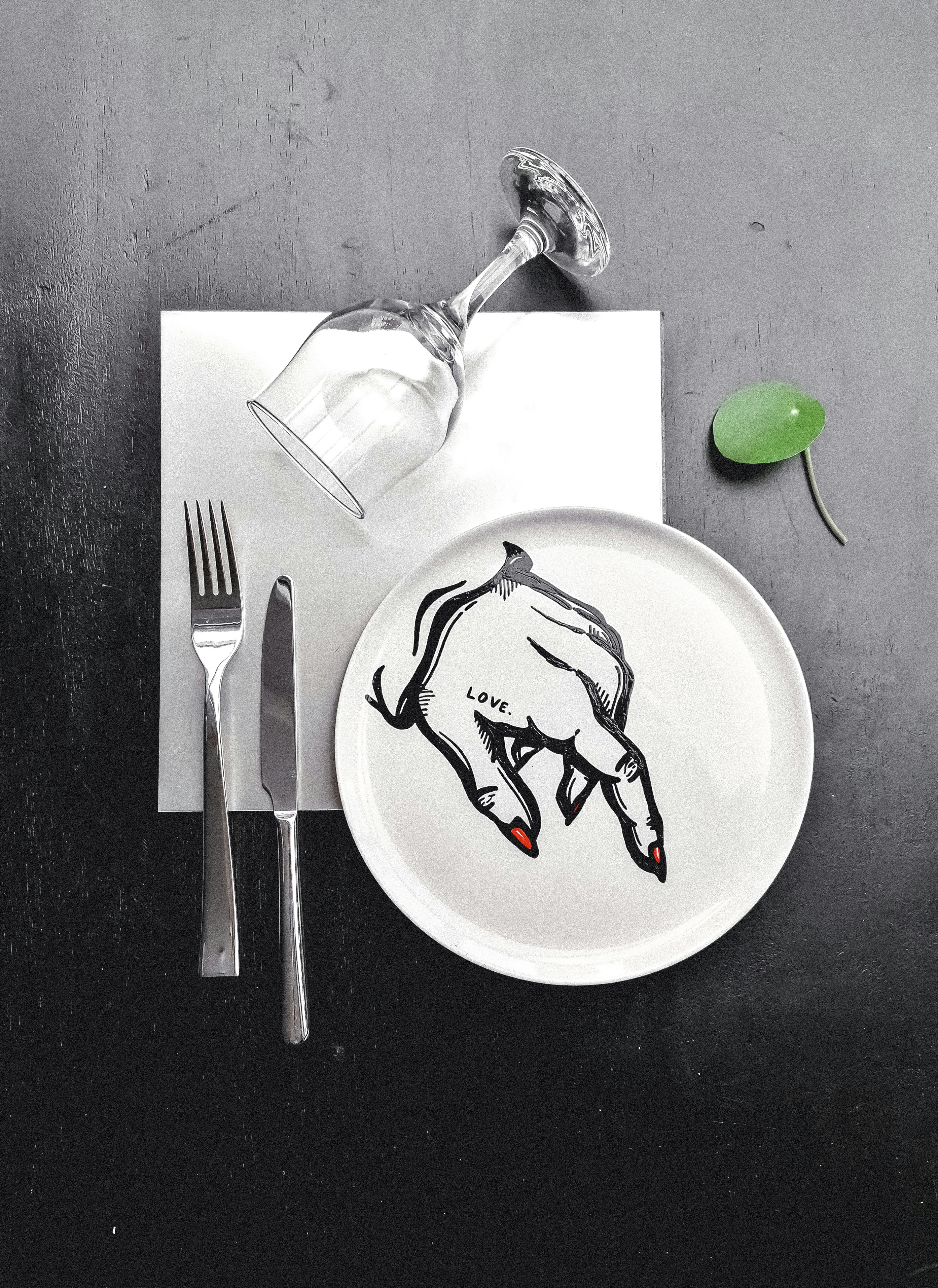 Still-life photograph of a plate bearing a black-ink illustration of a hand with the word LOVE, set on a dark tabletop. A nearby overturned wine glass, napkin, cutlery, and a single leaf complete the compact scene.