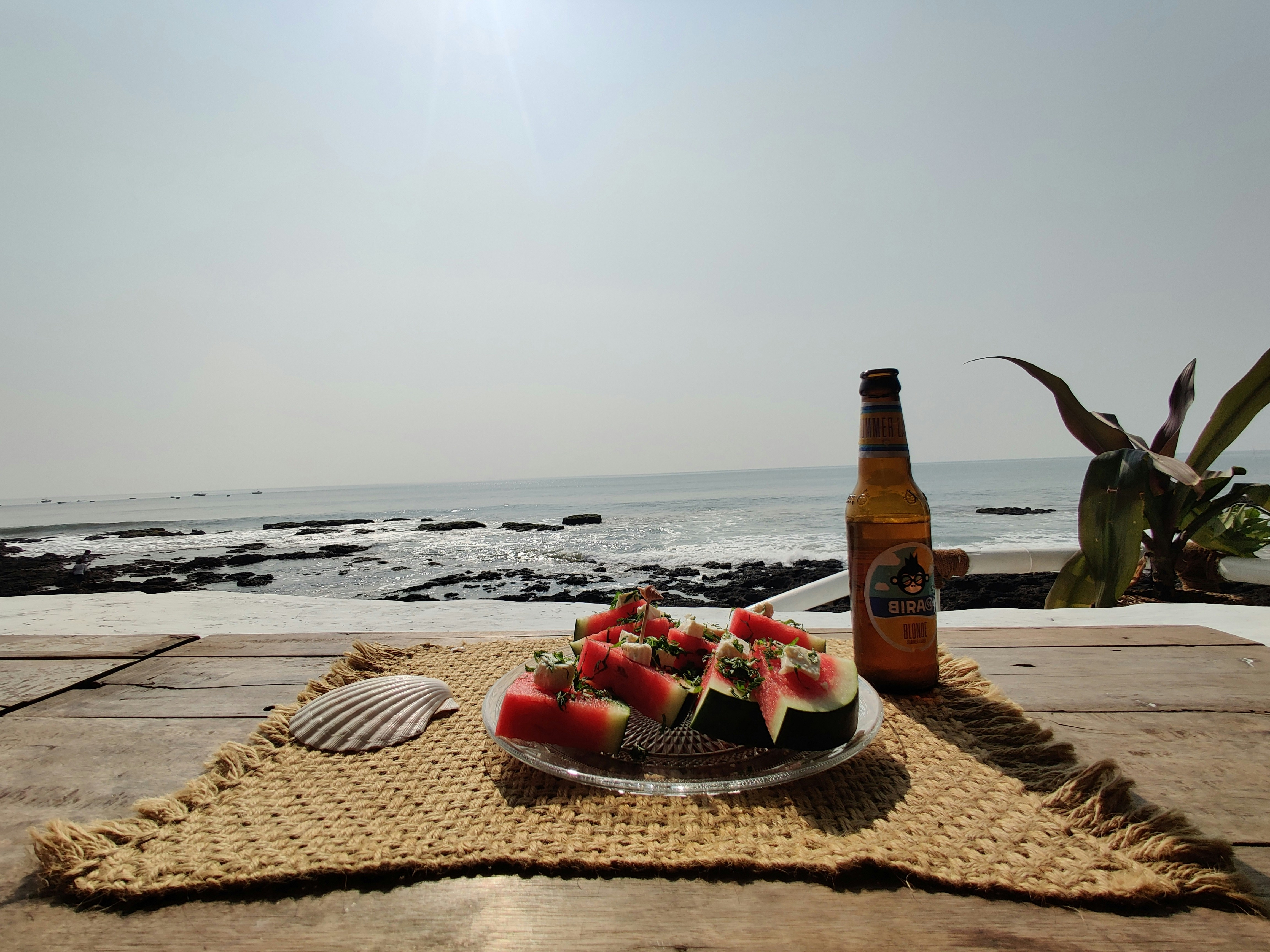 Sunlit seaside table with watermelon wedges on a woven mat and a beer bottle, overlooking the calm ocean.