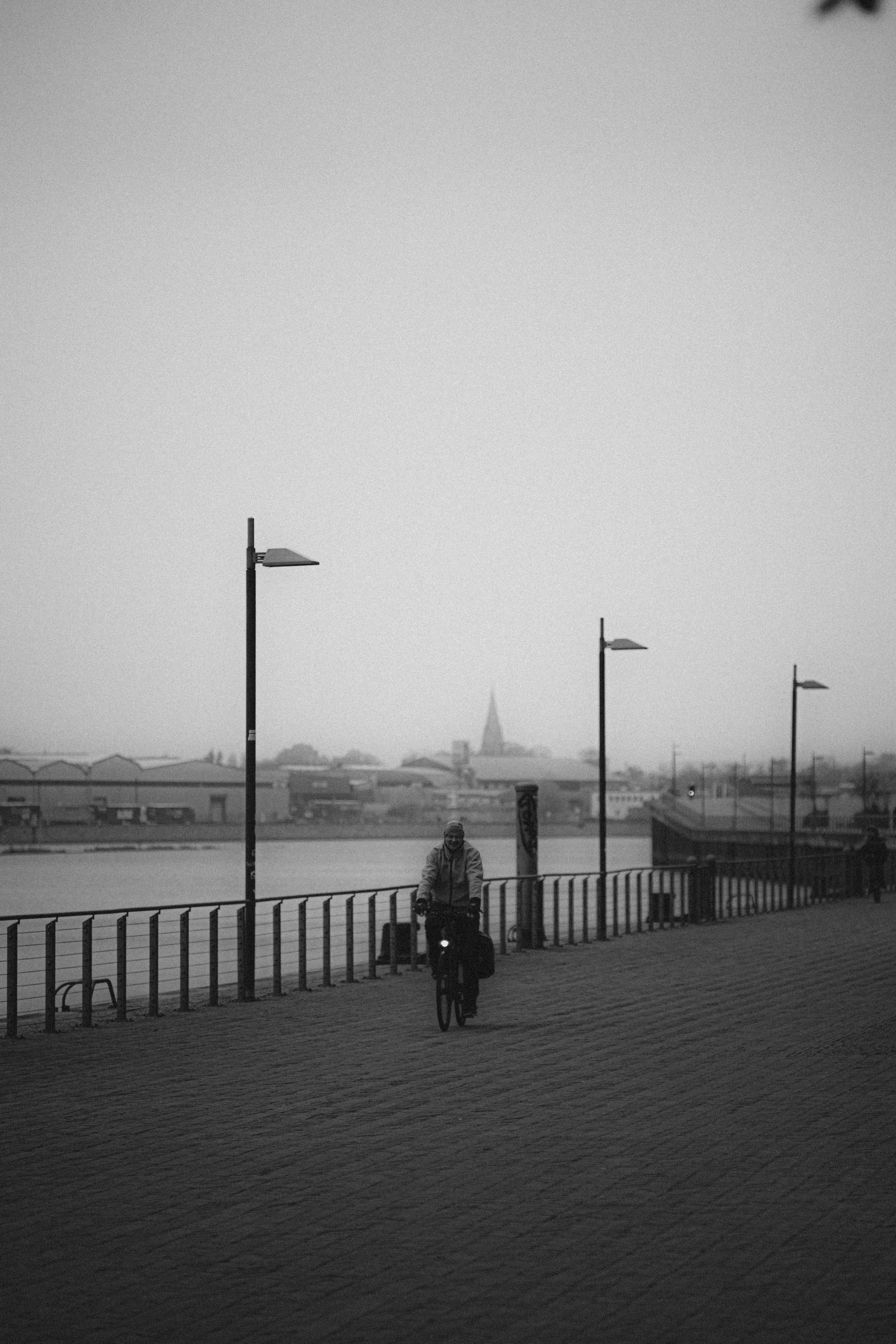 Cyclist traversing a foggy waterfront promenade, with distant buildings partially obscured by mist. The atmosphere evokes a sense of solitude.