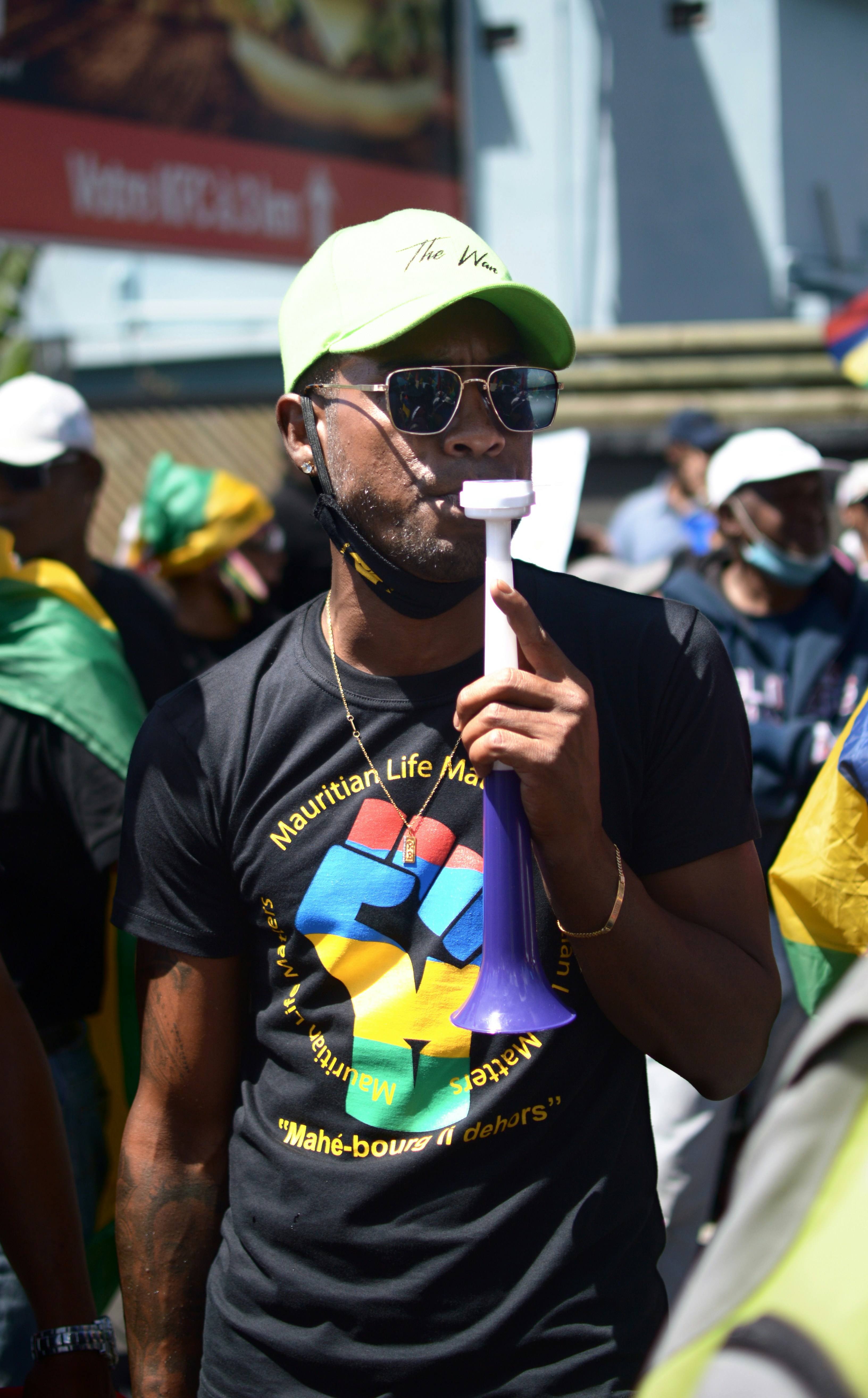 A man wearing sunglasses and a vibrant hat plays a trumpet during a lively street celebration, showcasing the colors and energy of Mauritian culture.