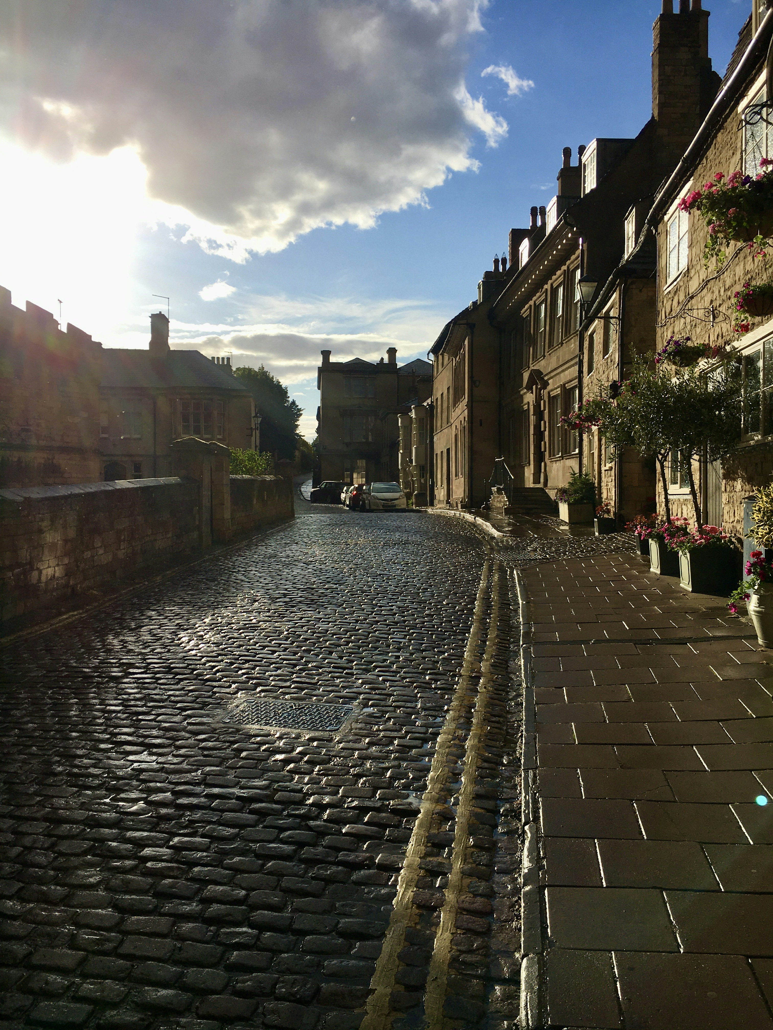 Sunlit cobblestone street flanked by historic stone buildings under a partly cloudy sky.