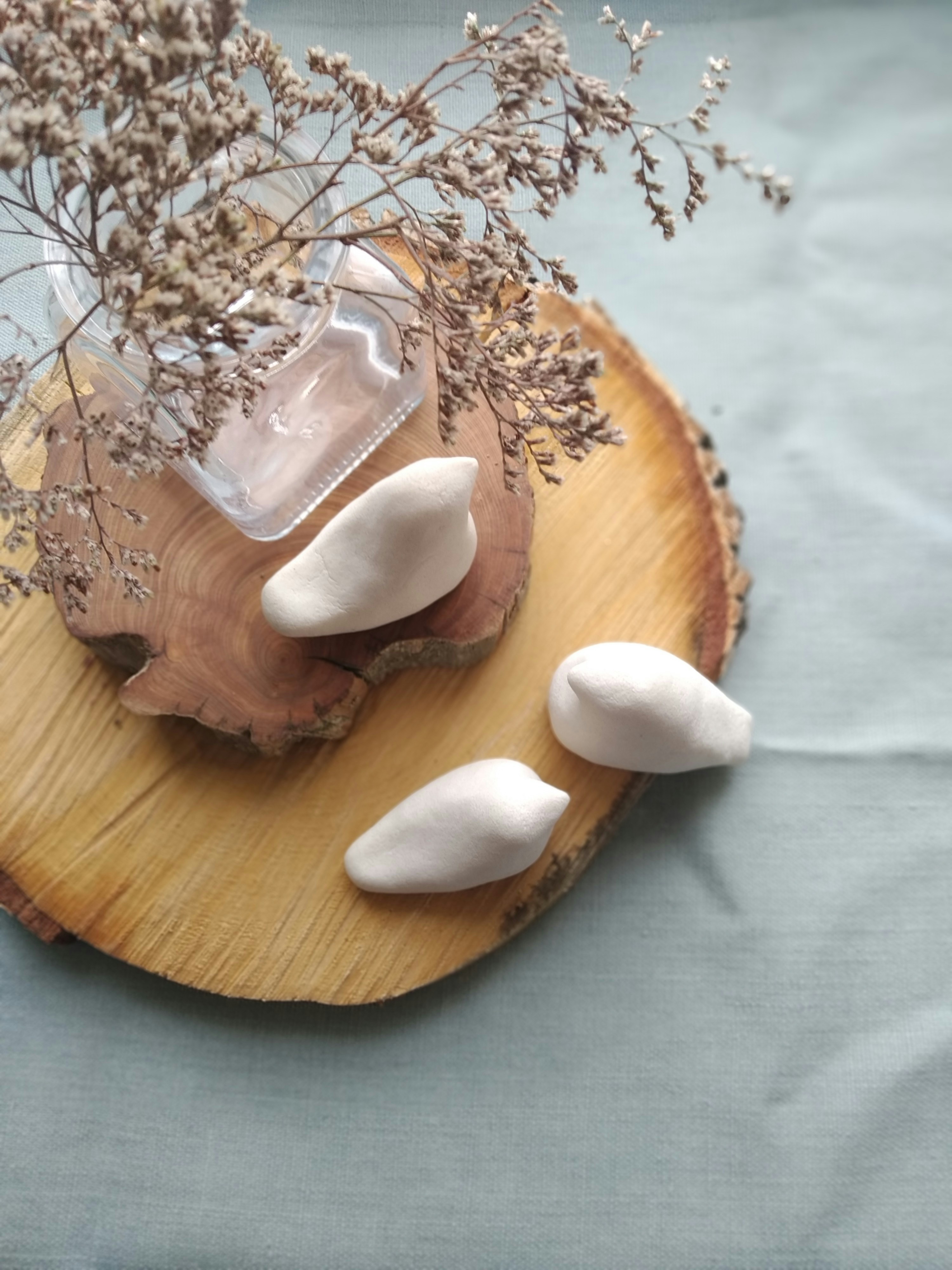 Delicate dried flowers in a glass container atop a wooden slice, accompanied by three smooth white stones, creating a serene and minimalist composition.
