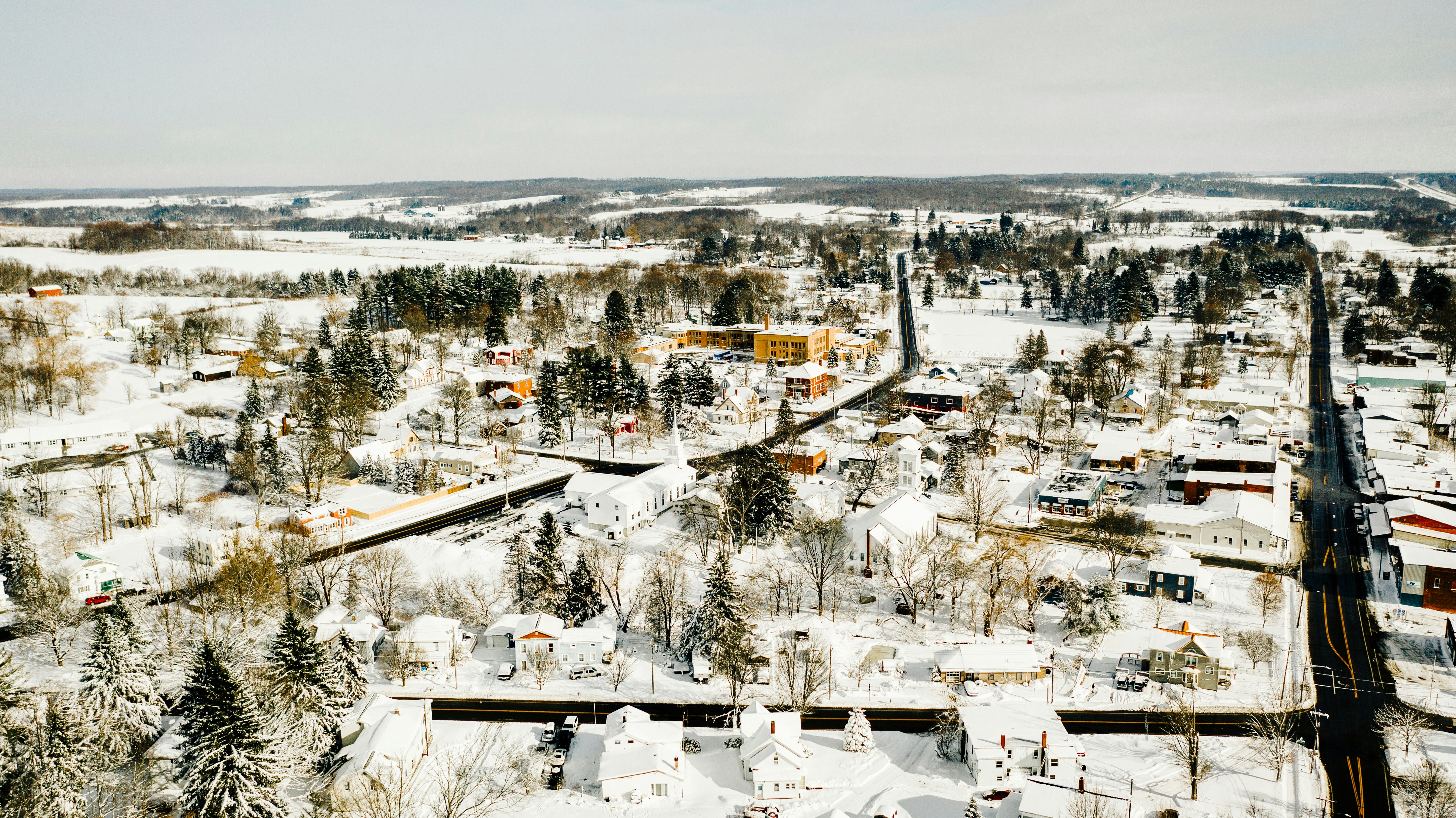 Aerial view of a quaint village blanketed in fresh snow, showcasing rooftops and tree-lined streets. The landscape is serene, with a gentle contrast between the white snow and the colorful buildings.