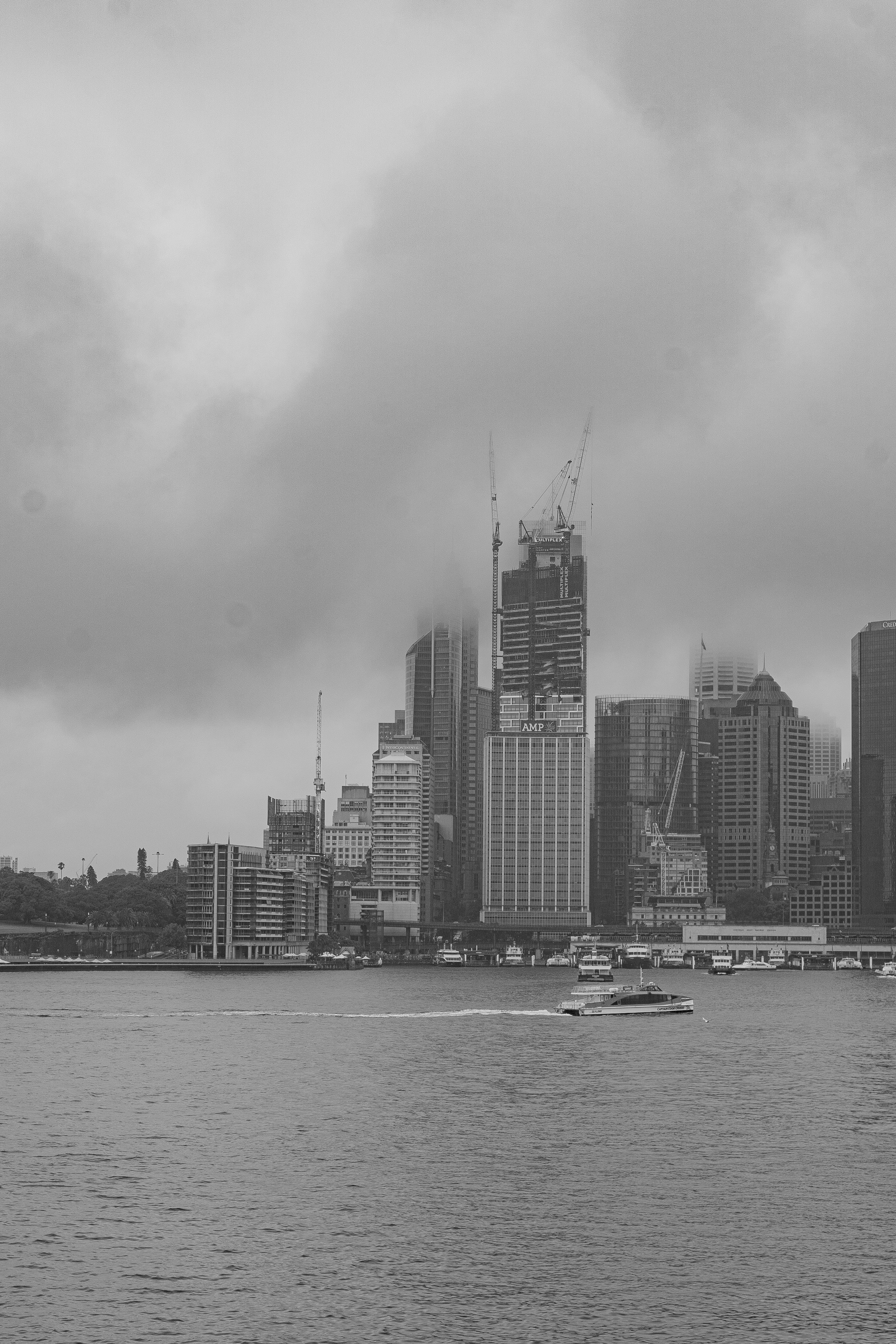 Monochrome cityscape featuring skyscrapers partially shrouded by low-hanging clouds, with a boat navigating the water in the foreground.