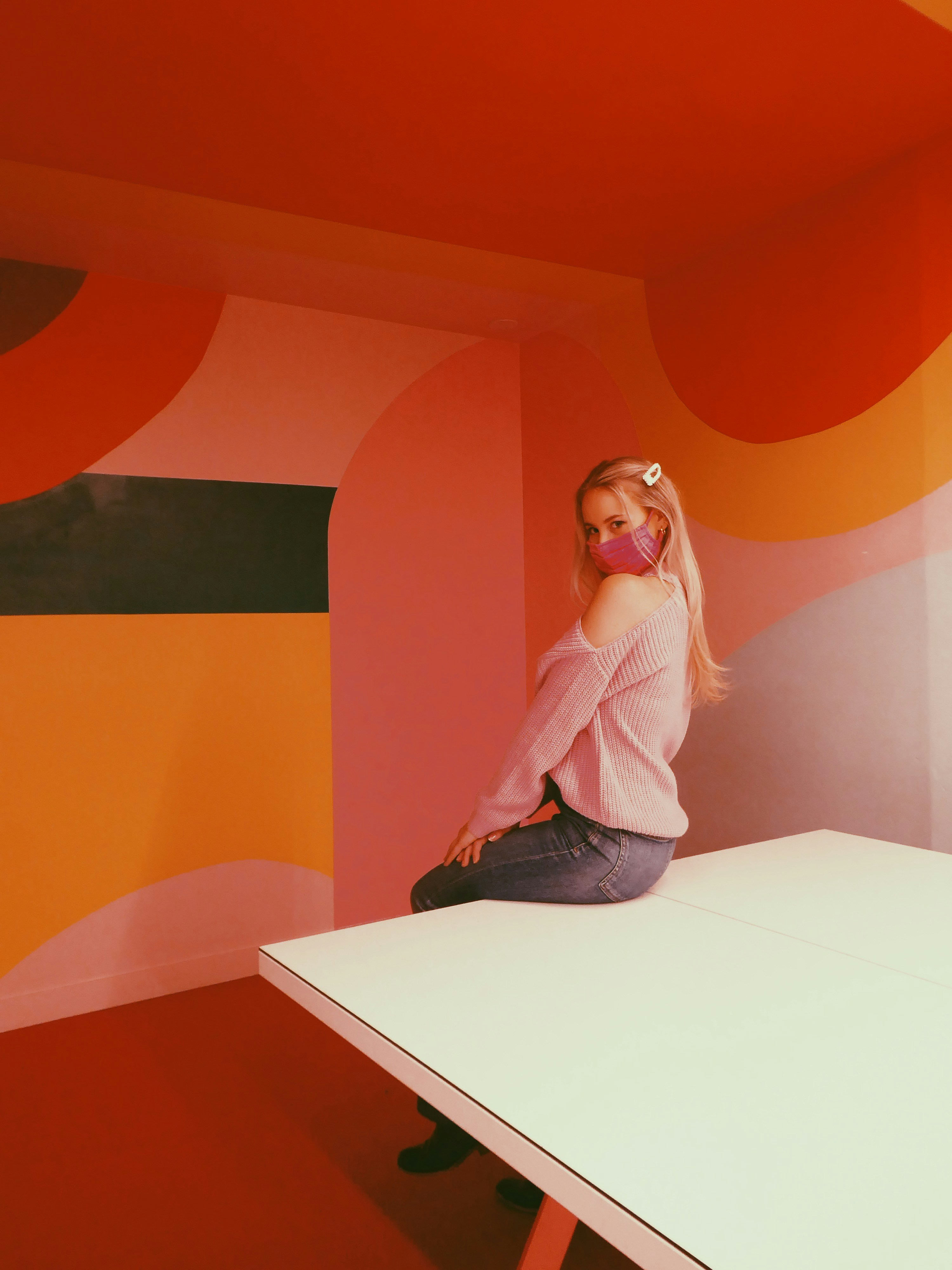 A young woman poses playfully on a table amidst a vibrant, abstract mural of shapes and colors. Her relaxed demeanor contrasts with the lively background.