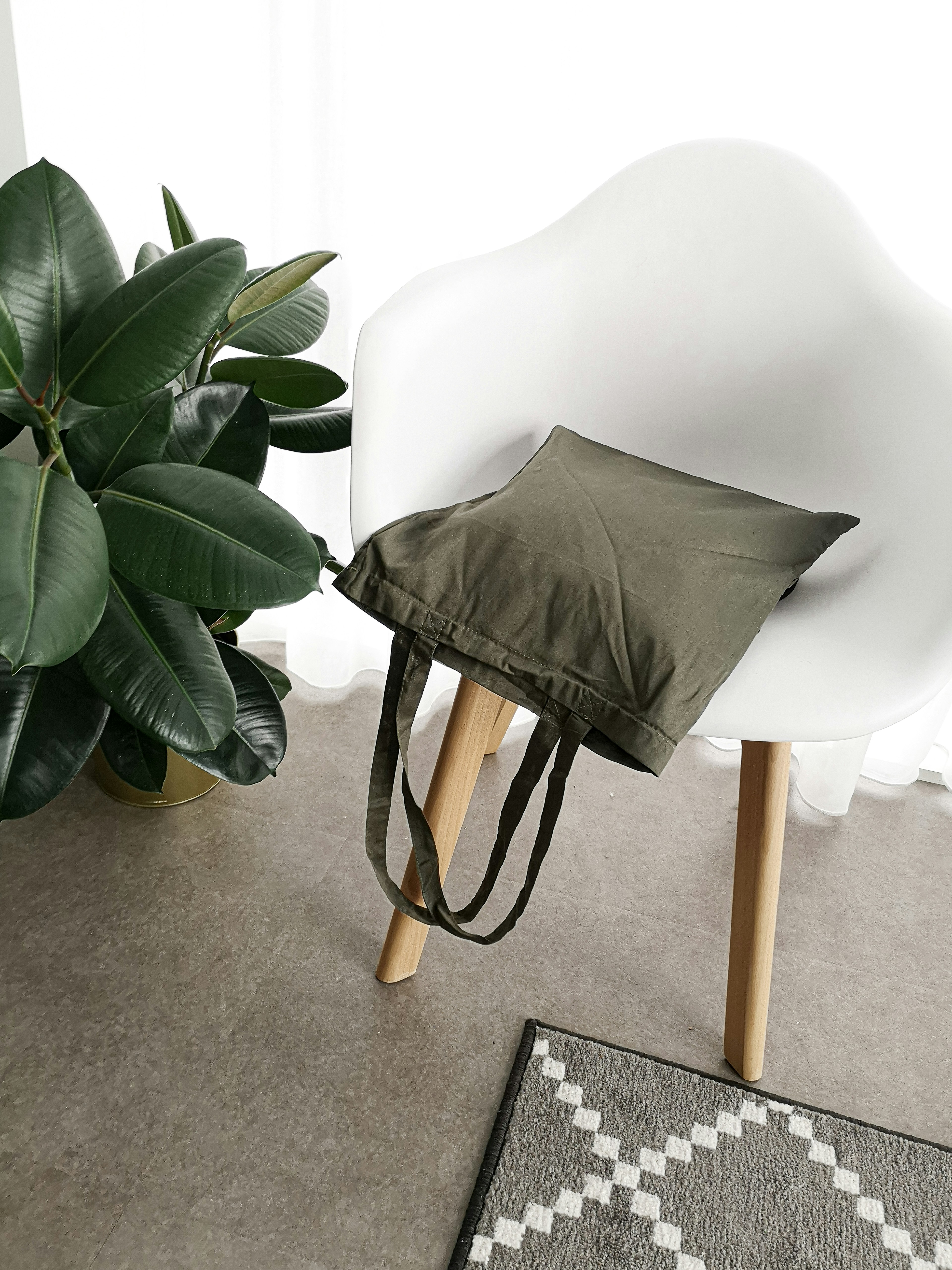 A minimalist scene featuring an olive green tote resting on a sleek white chair beside a lush potted plant, set against a bright and airy backdrop.