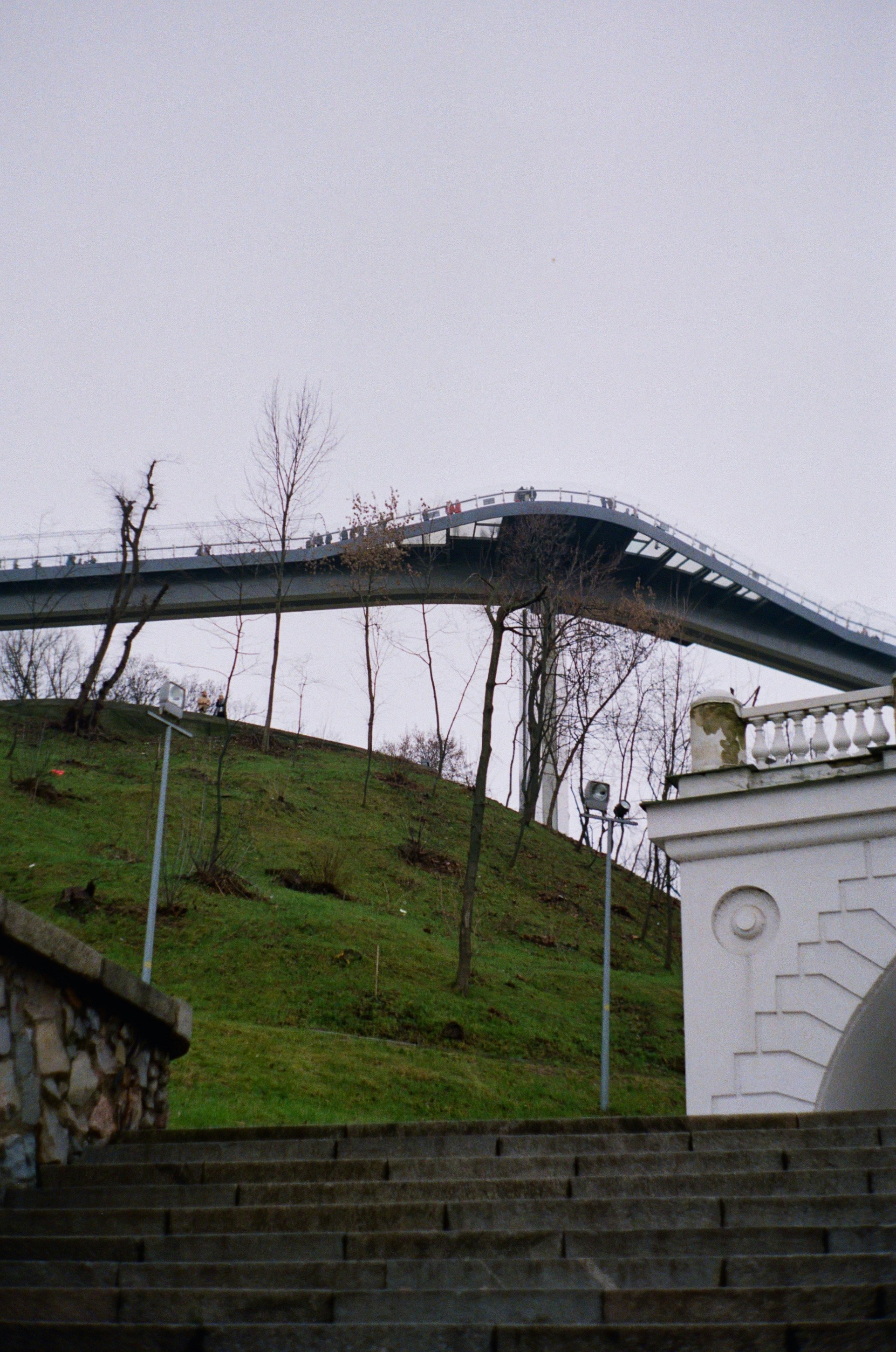 A modern pedestrian bridge gracefully arches over a hillside, surrounded by bare trees and a hint of spring greenery. The scene captures an intriguing blend of architecture and nature.