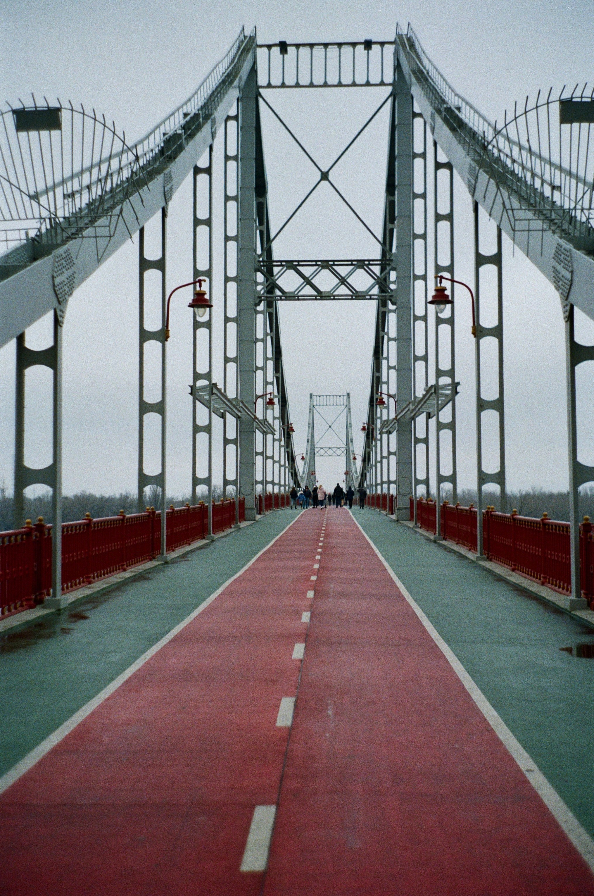 A vibrant red pathway stretches across a modern suspension bridge, flanked by intricate steel structures and soft gray skies. Pedestrians traverse the bridge, adding life to the scene.