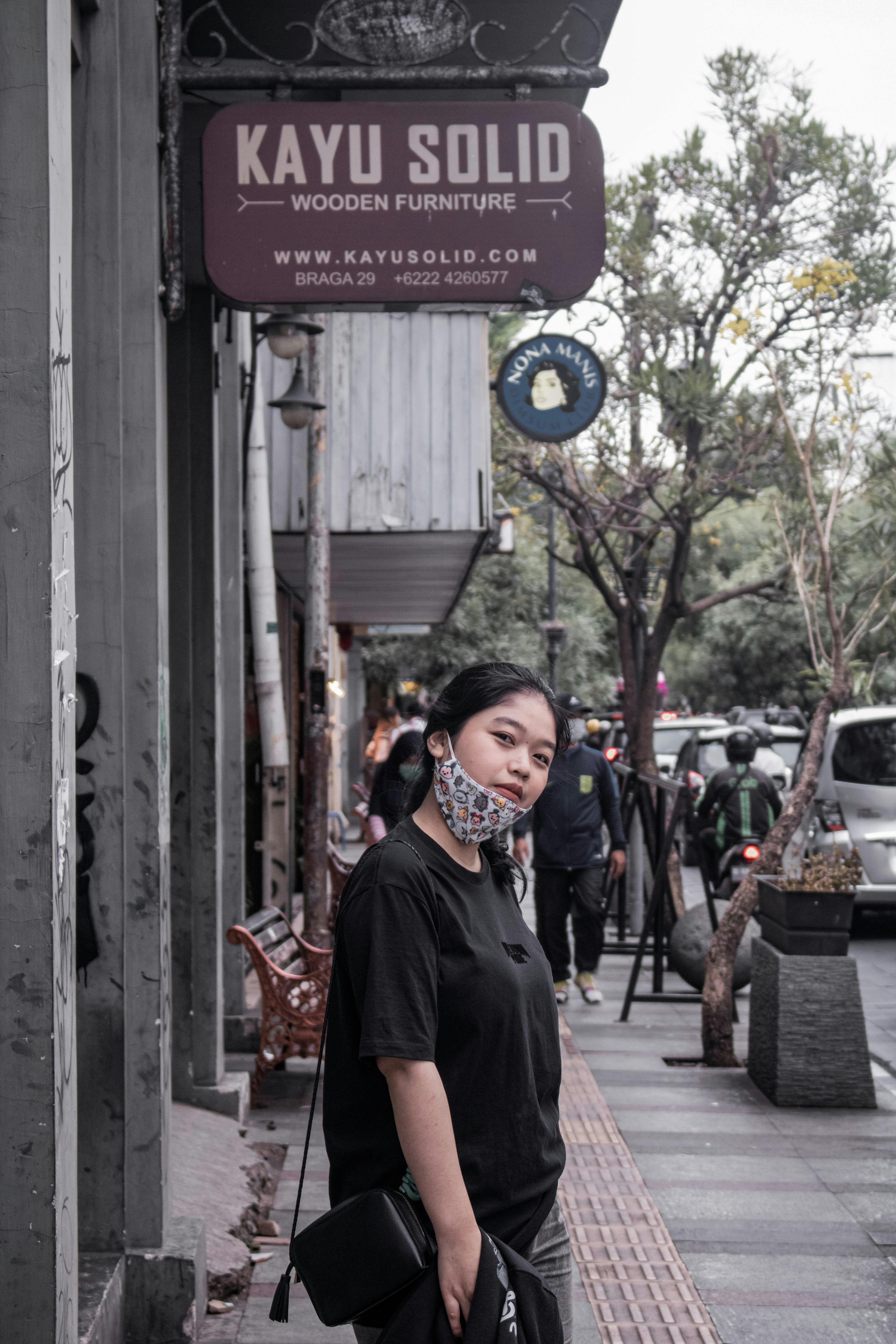 A young woman poses in a casual black outfit on a lively street, showcasing a vibrant urban atmosphere with shops and greenery in the background.