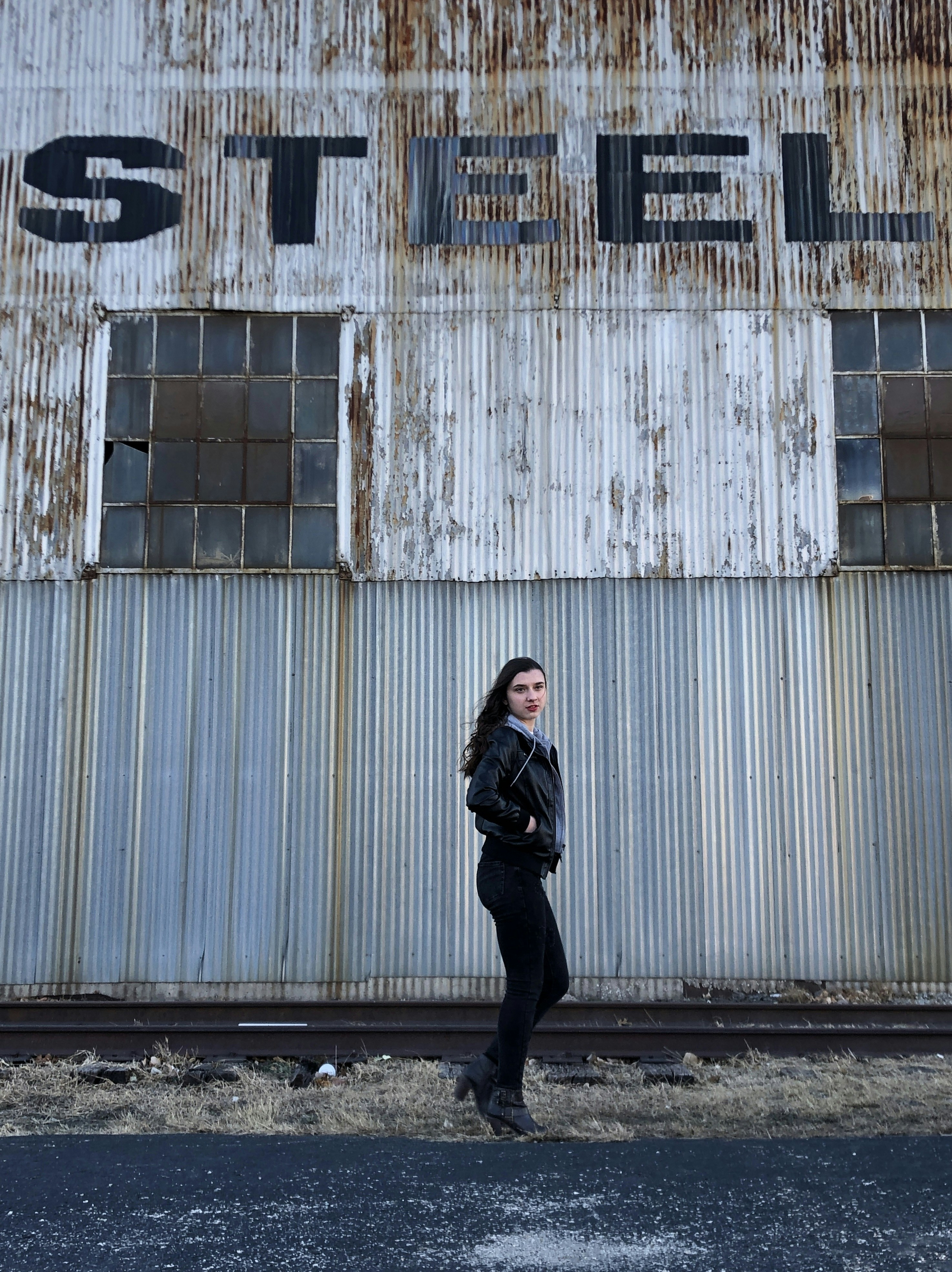 A young woman poses confidently in front of a weathered steel wall, embodying urban resilience and style.