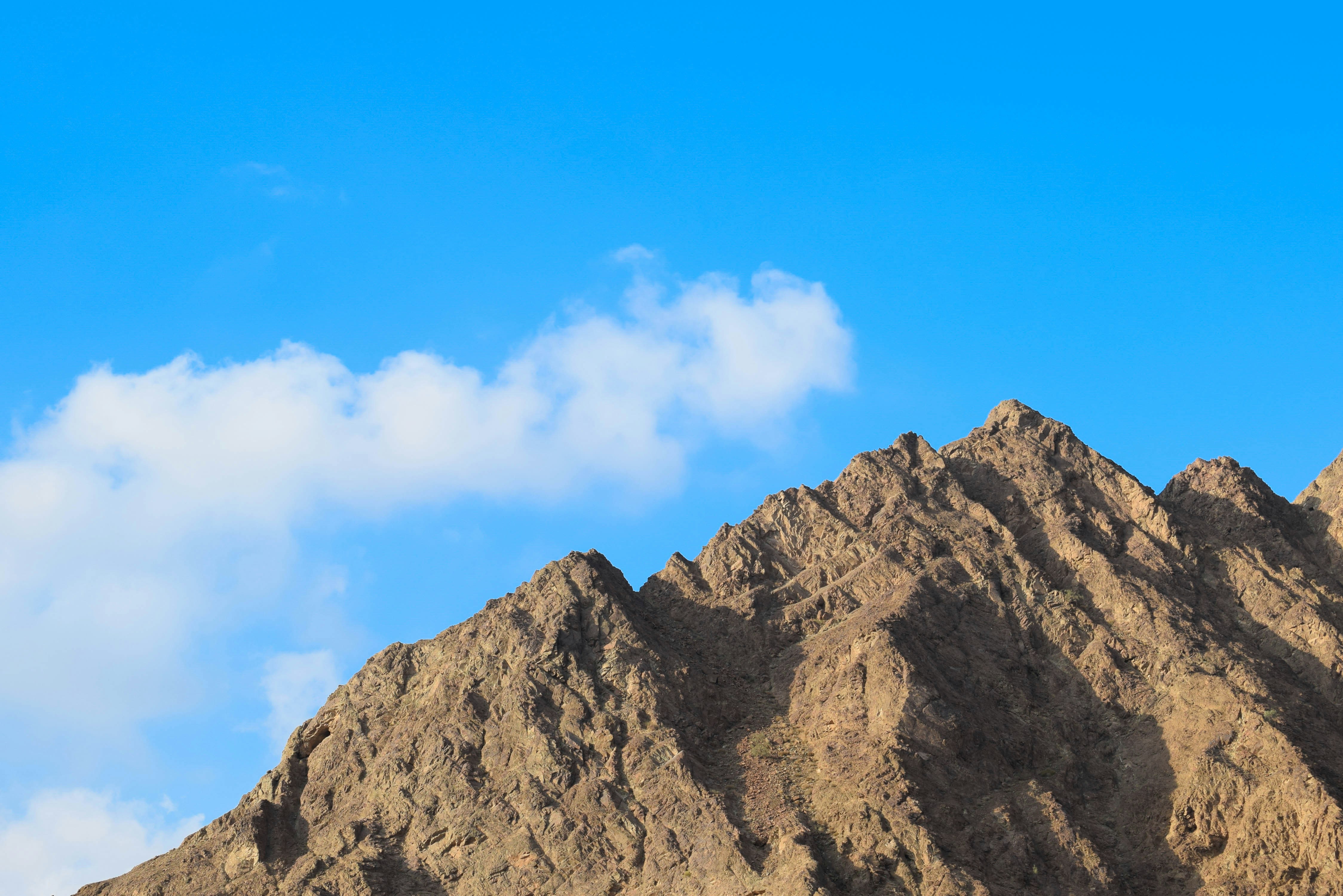 Jagged mountain ridges rise sharply against a clear blue sky, with soft clouds drifting by. The scene captures the raw beauty of nature's rugged landscape.