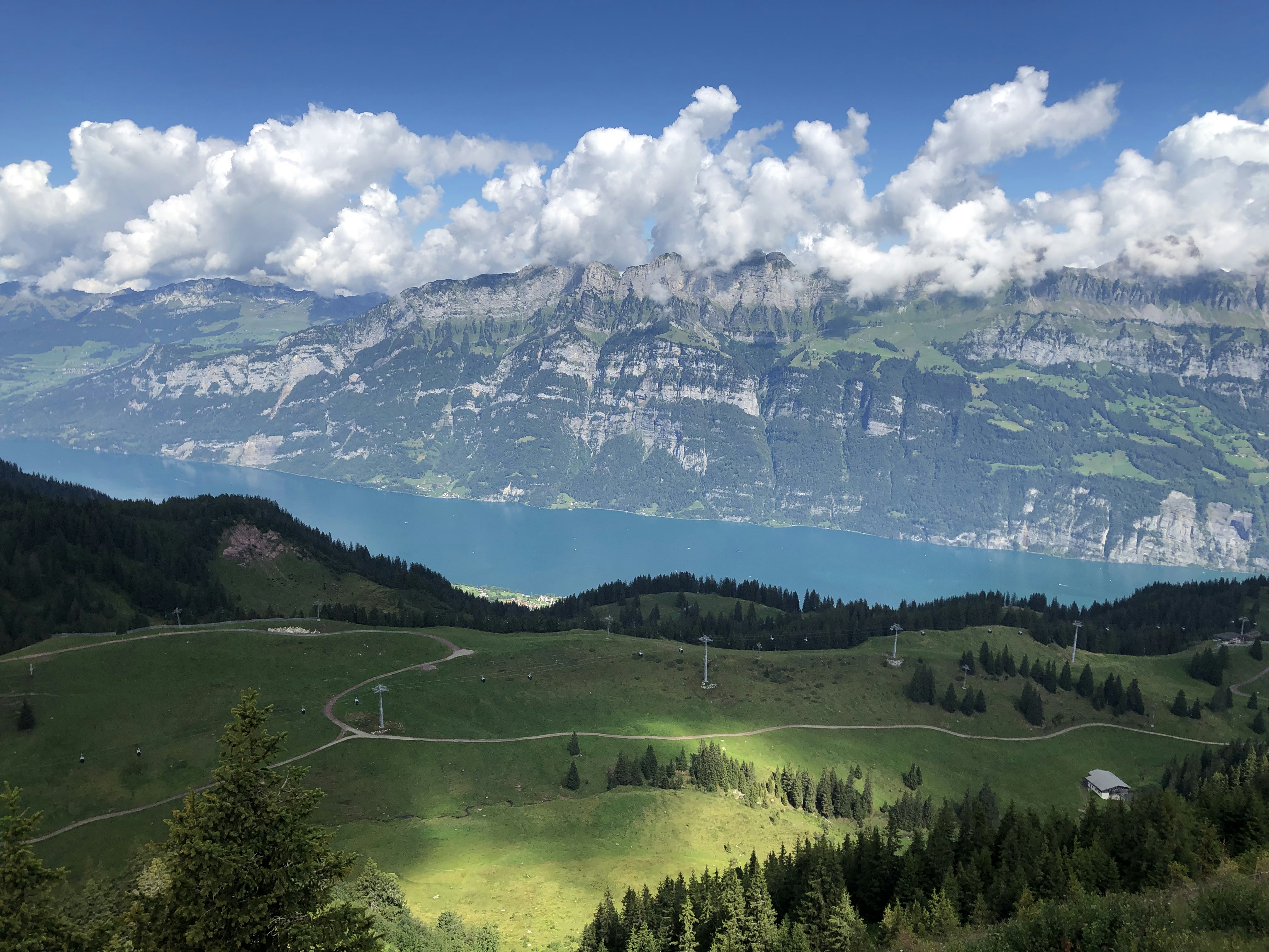 Vast green meadows and towering mountains reflect in the serene waters of a lake under a dynamic sky filled with clouds.