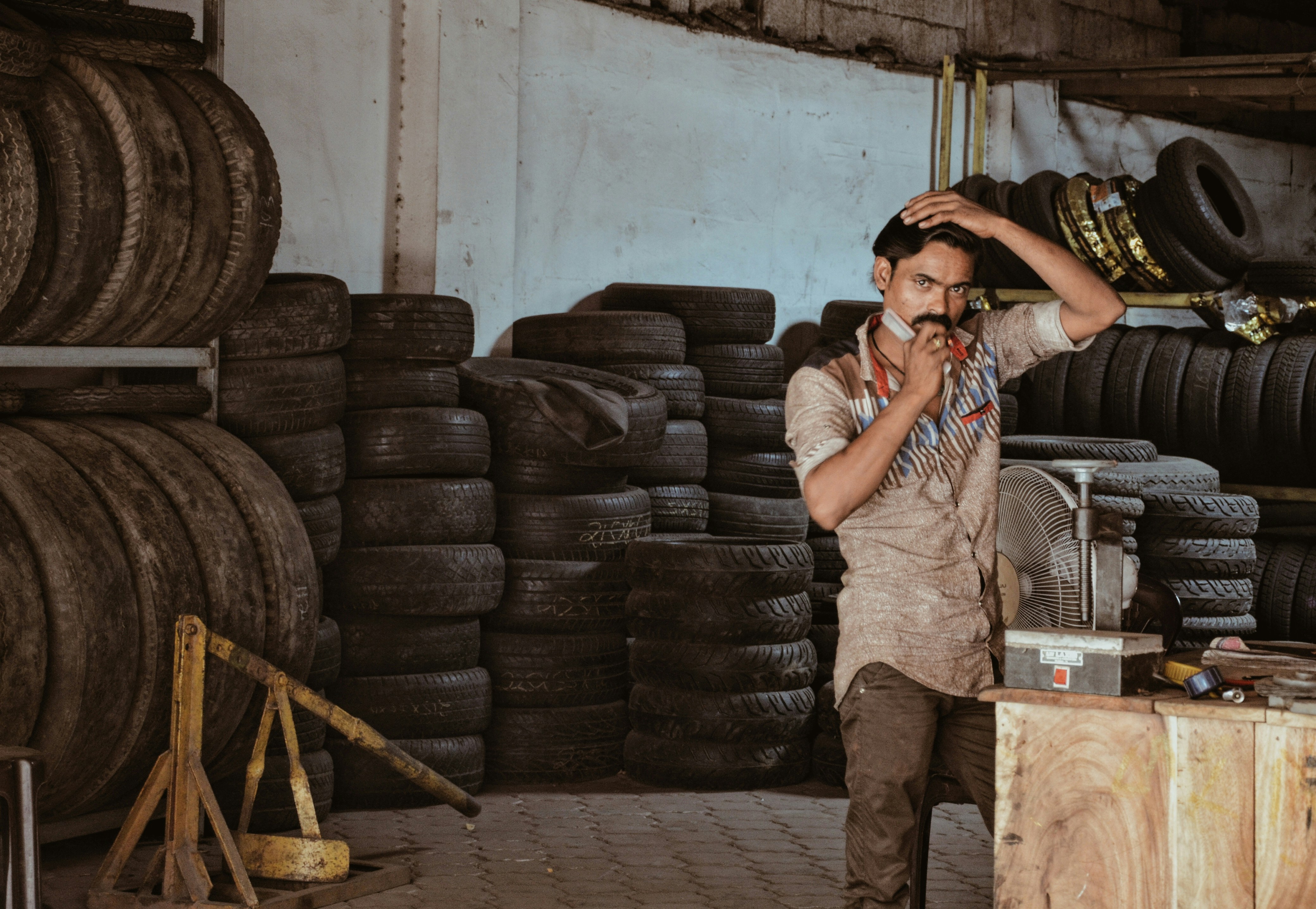 A mechanic stands pensively in a tire shop, surrounded by stacks of used tires, contemplating his next move. The setting highlights the gritty atmosphere of the workshop.