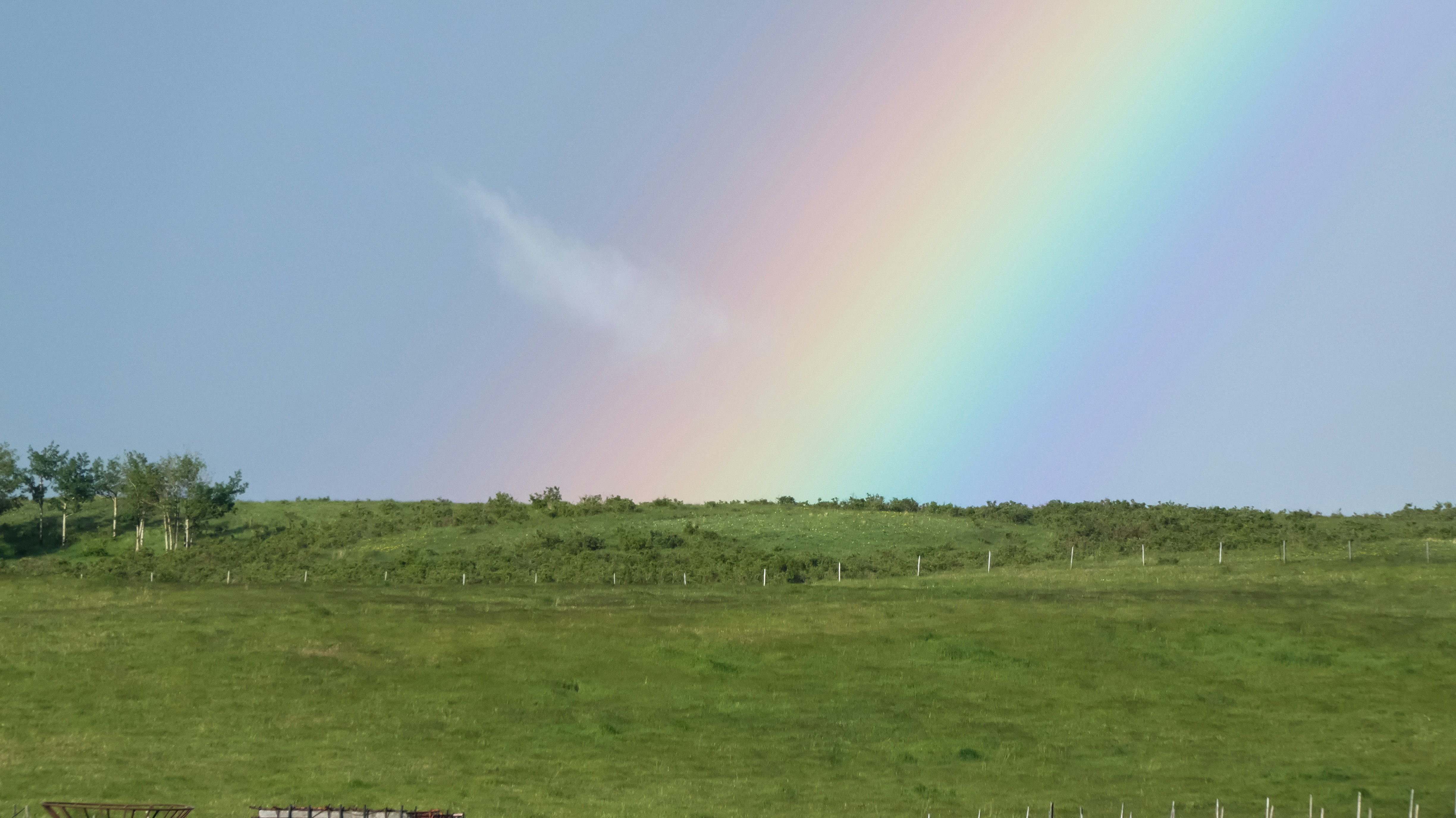 Pastureland with a gentle slope and fence posts under a blue sky as a vivid rainbow arches across the horizon.